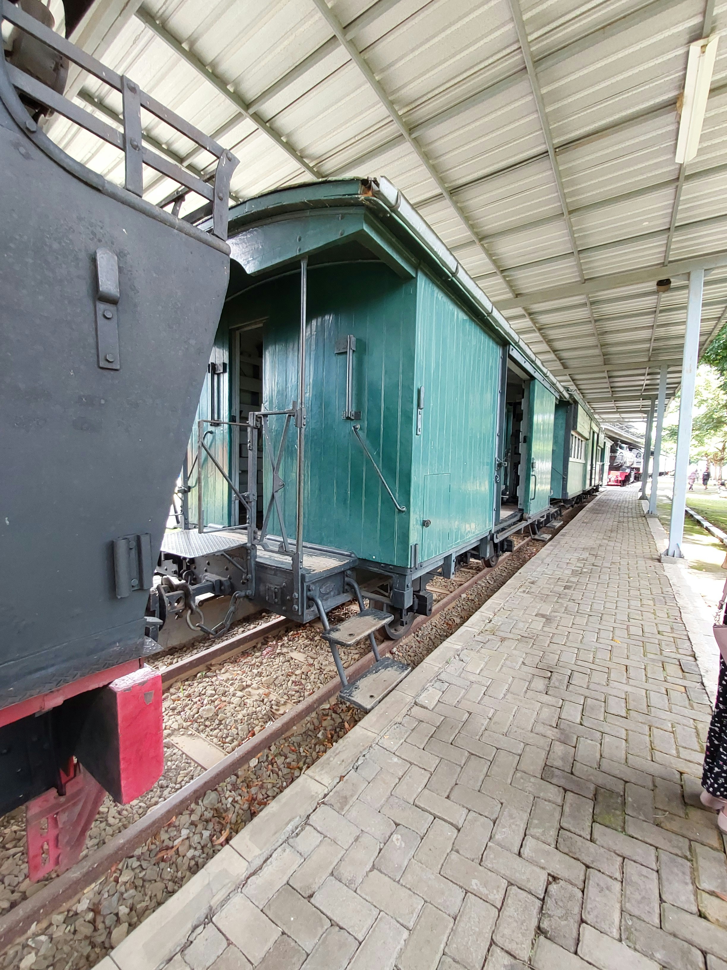 A vintage green caboose sits on a weathered track beneath a metal canopy, stretching along a sunlit platform. The long row of railcars and the station's repeating rails create a strong sense of depth.