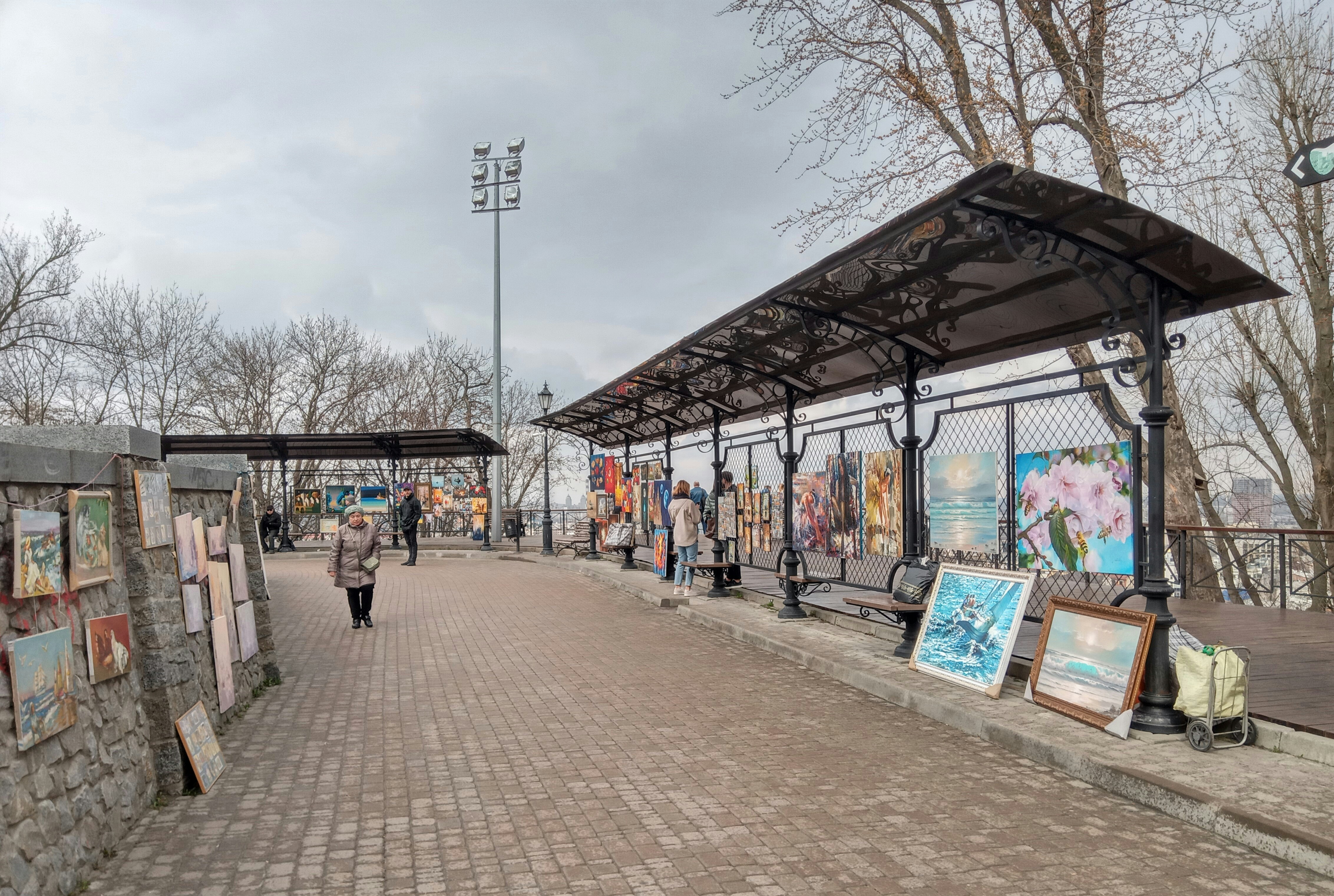 Riverside art promenade with framed paintings displayed along a cobblestone path beneath a metal canopy. Visitors browse the artwork on a gray, overcast day.