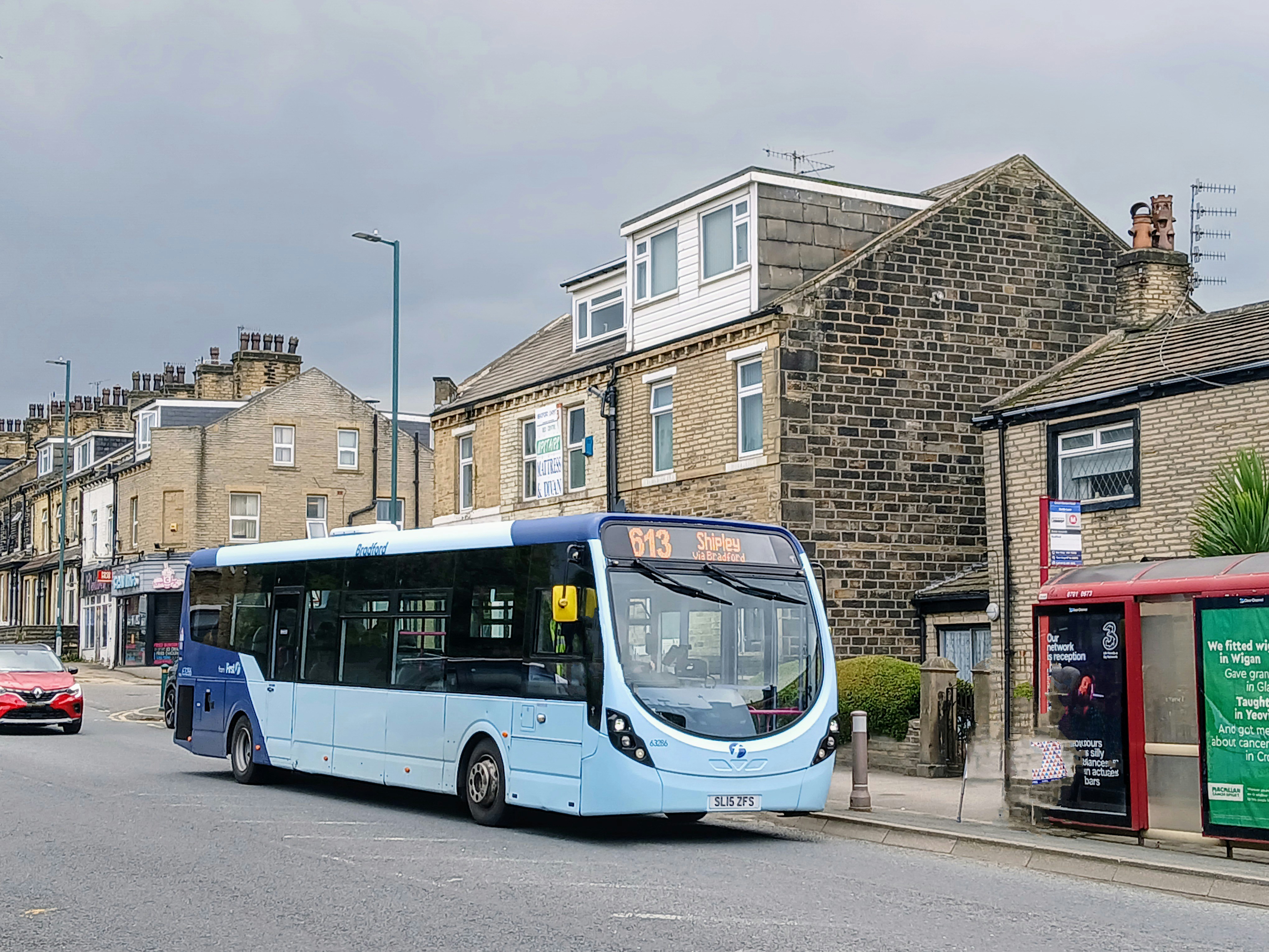 City bus navigating a quiet street, flanked by traditional buildings and a bus stop. The scene captures daily life in an urban setting.