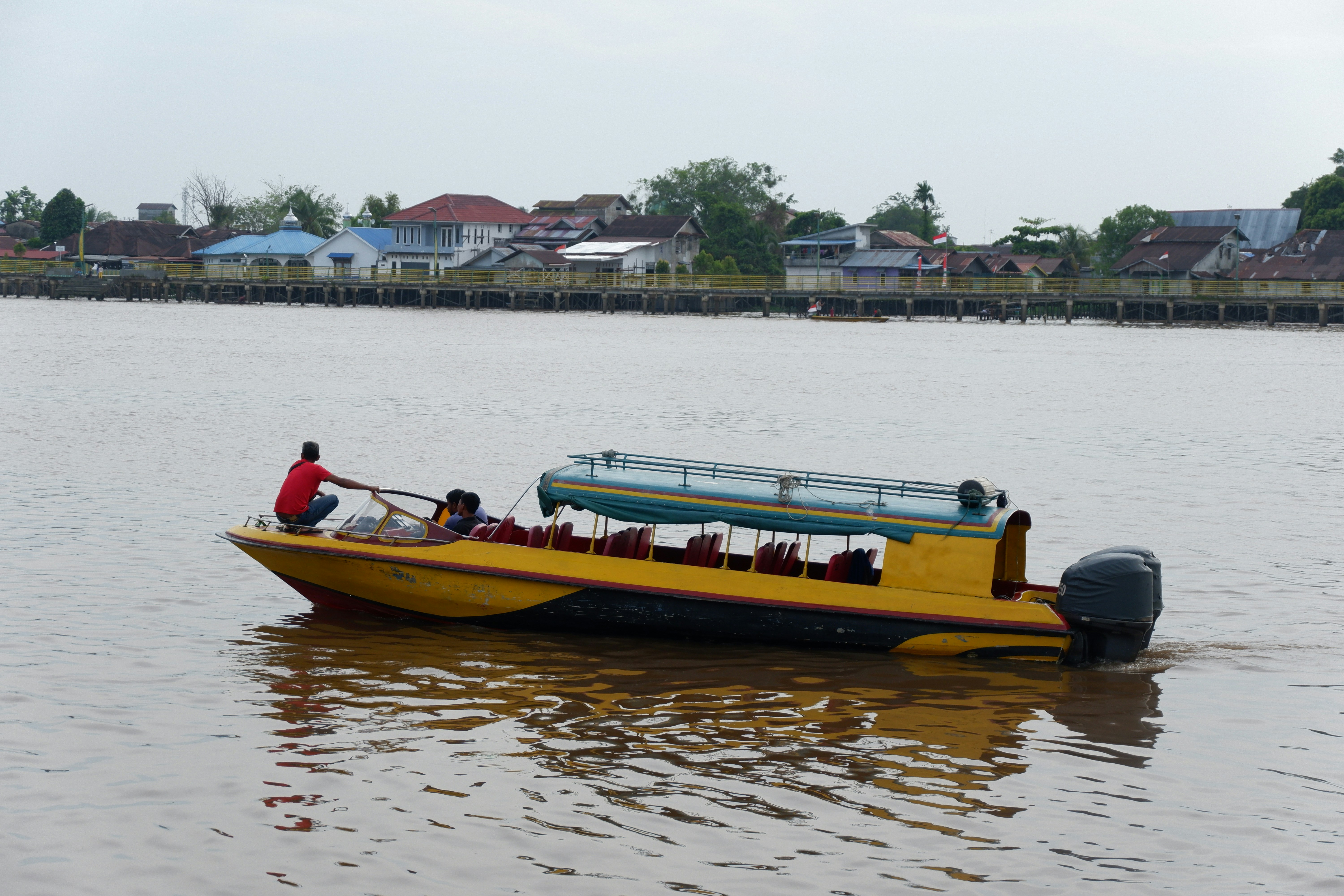 Yellow boat with a canopy glides across a calm river, with a distant village lining the opposite bank under a cloudy sky.