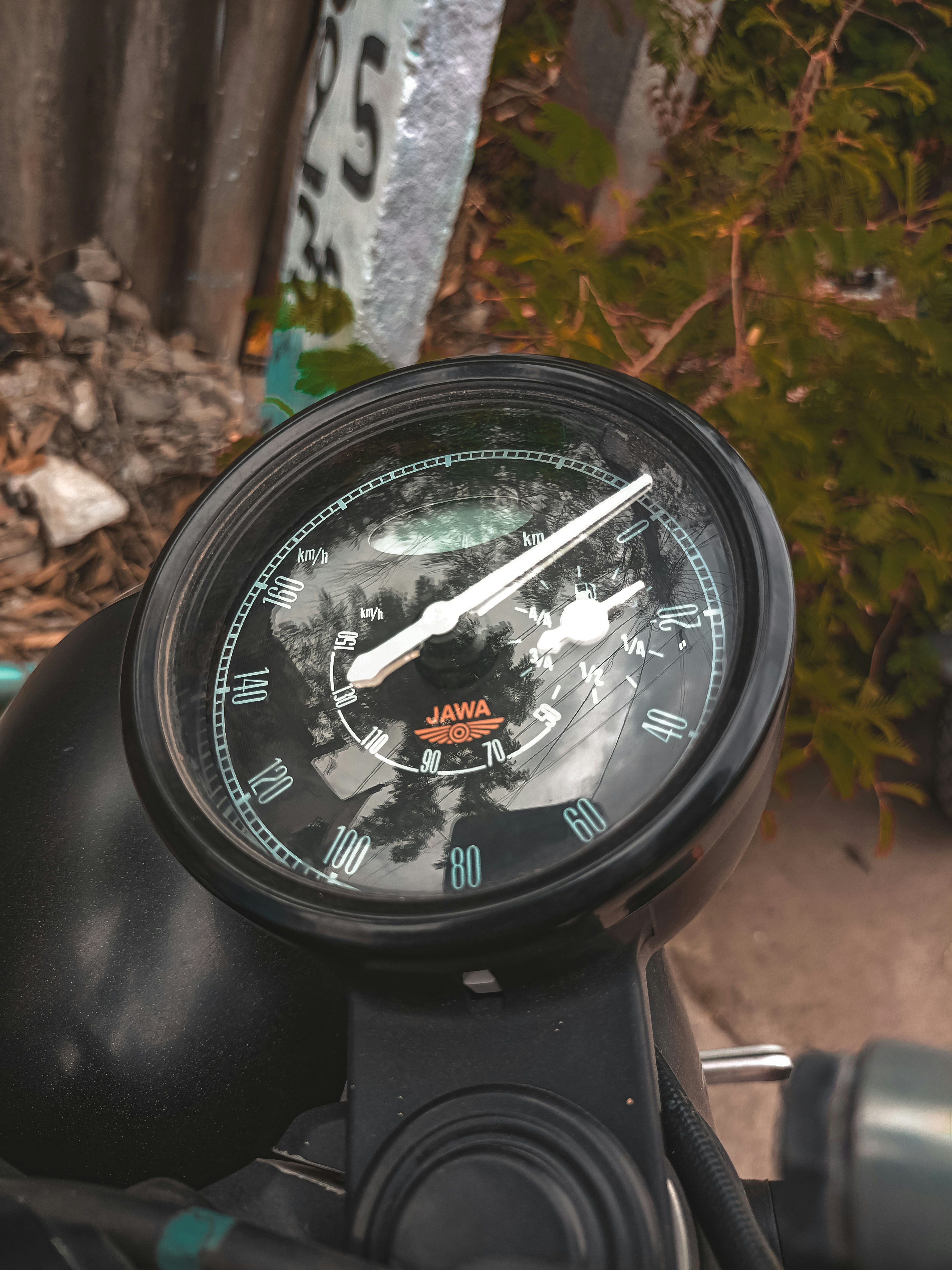 Close-up view of a round motorcycle speedometer with reflections and surrounding handlebars, outdoors with dirt and foliage in the background. The image appears to be a photo capture of a real object rather than an illustration.
