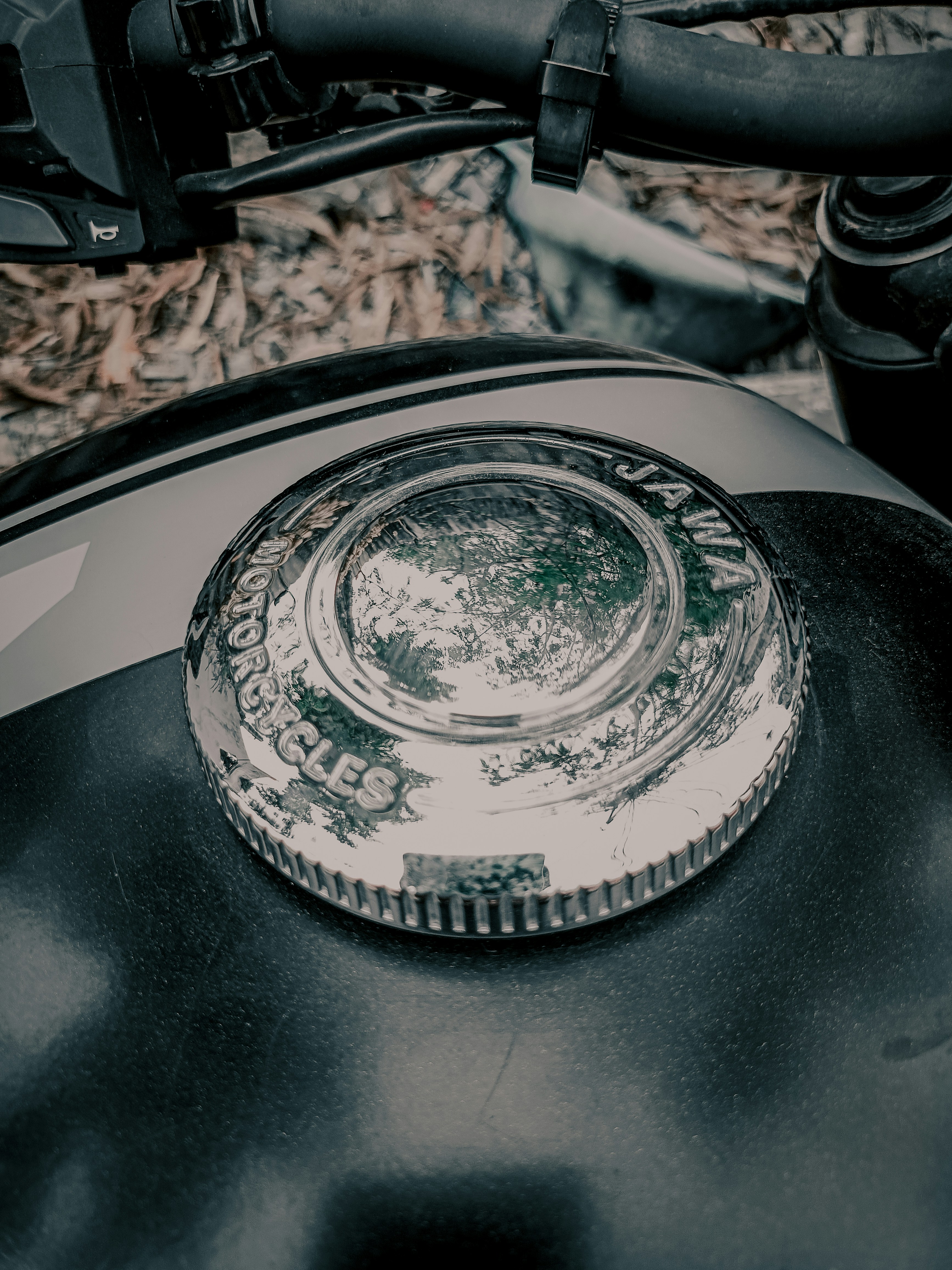 Close-up photograph of a chrome motorcycle fuel cap on a dark tank, with reflections and surrounding cables.