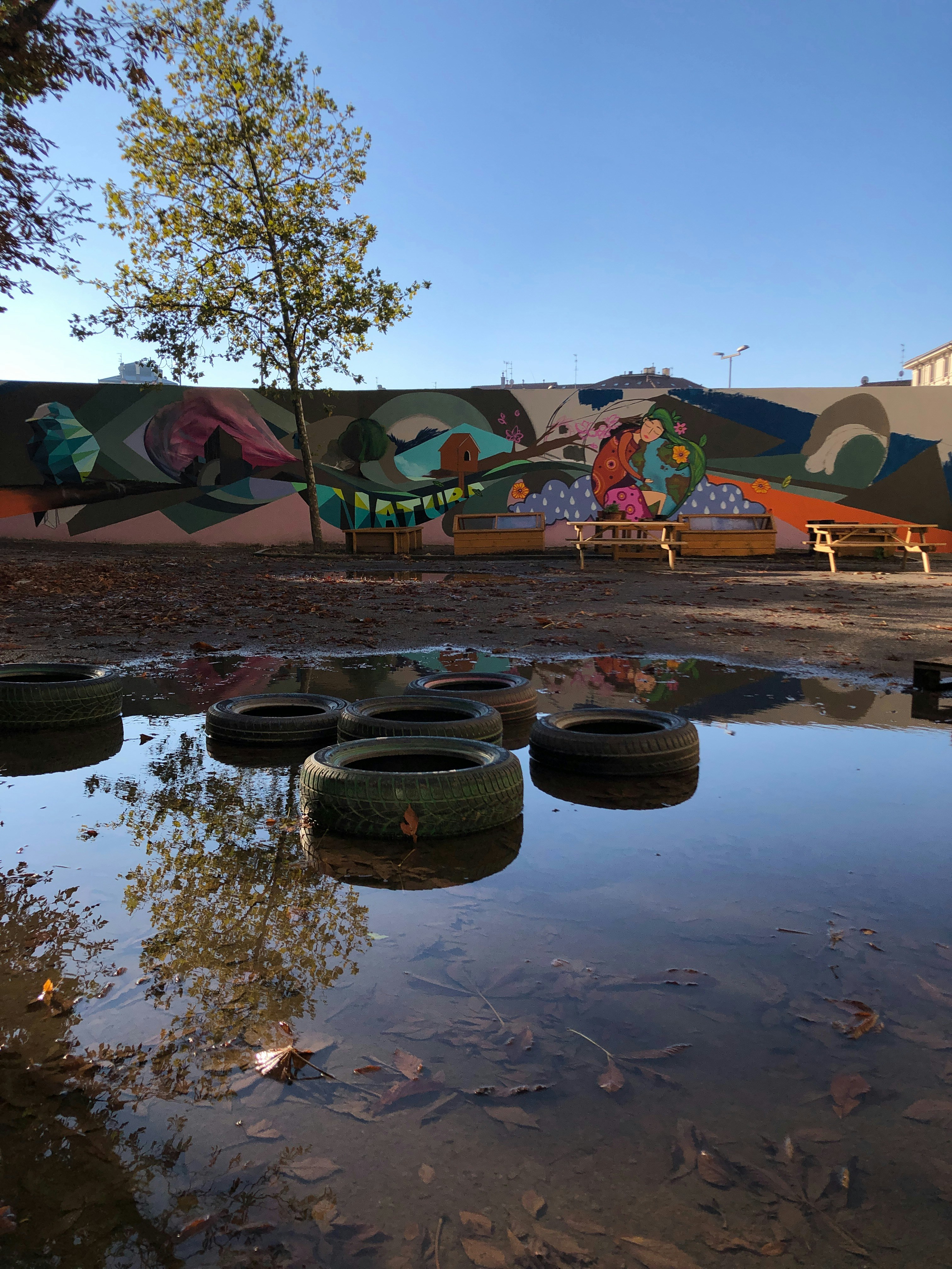 Colorful mural on urban wall mirrored in a puddle surrounded by old tires and autumn leaves, showcasing the intersection of nature and art.