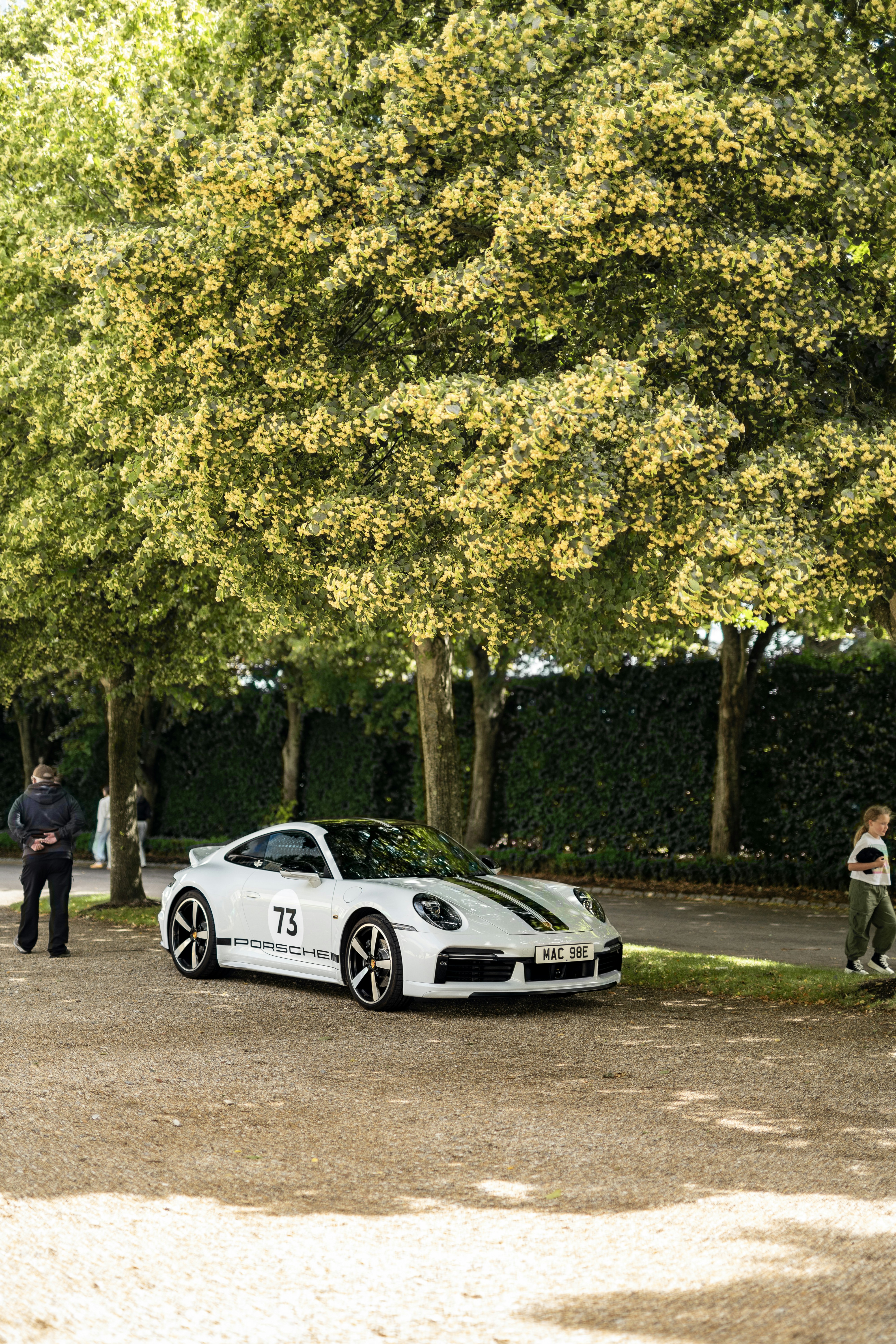 White Porsche 911 parked beneath a canopy of trees during a sunny day event