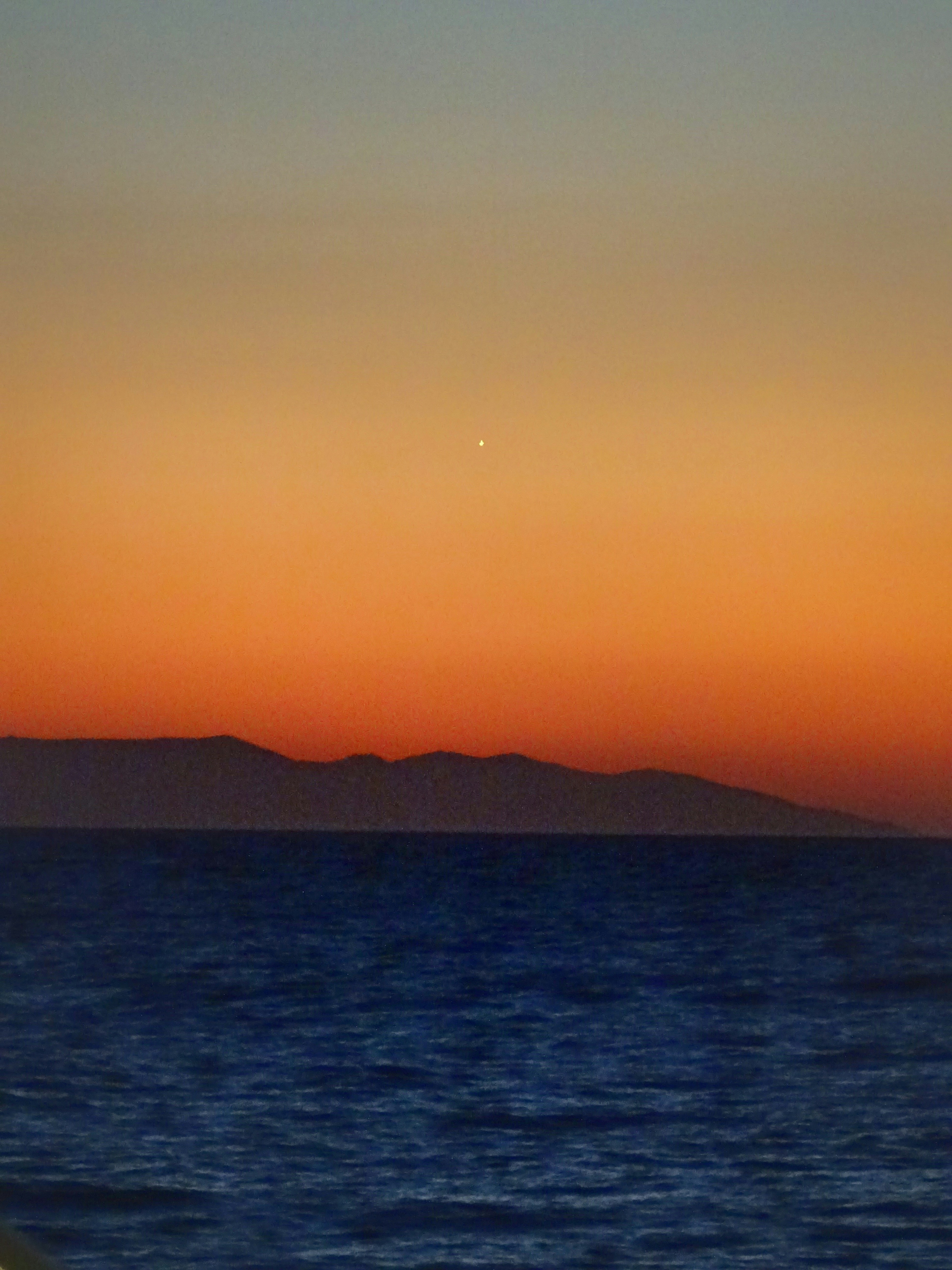 Photo of a sunset over the sea with a silhouetted landmass along the horizon. A solitary bright dot rests in the evening sky above calm waters.