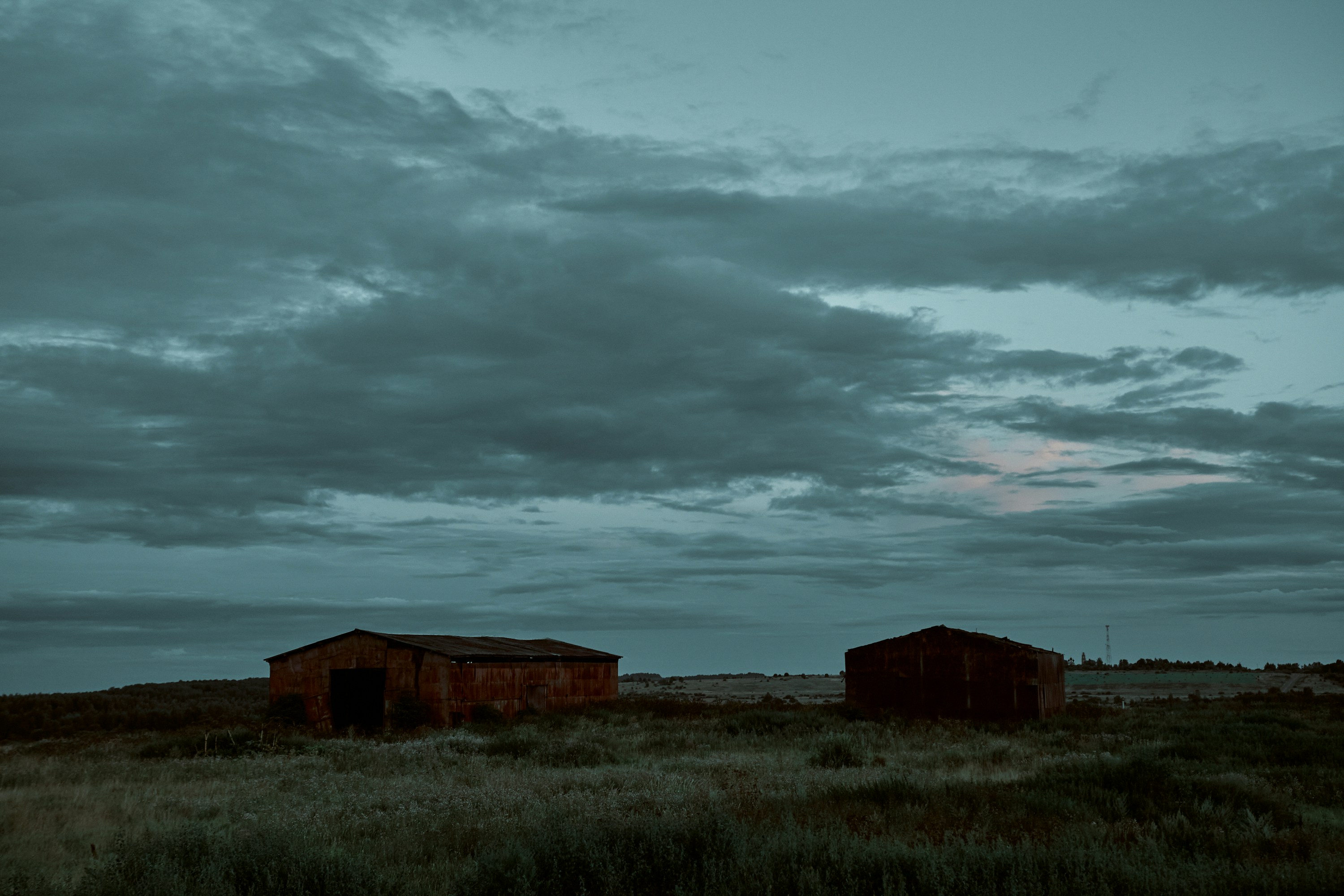 Two rustic buildings stand isolated under a cloud-heavy sky in a desolate landscape at dusk.