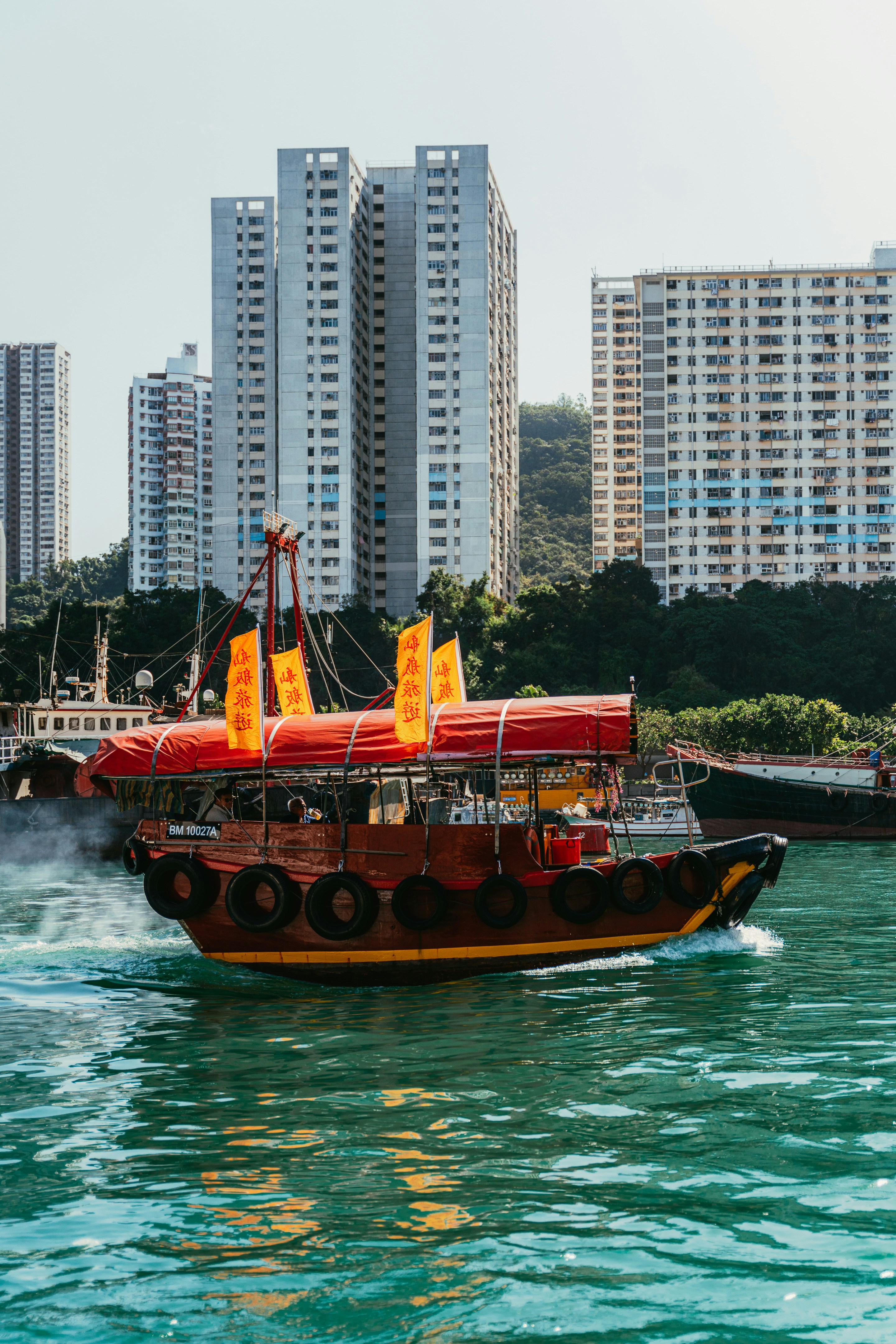 Traditional junk boat with vibrant sails glides past towering city buildings and lush greenery.