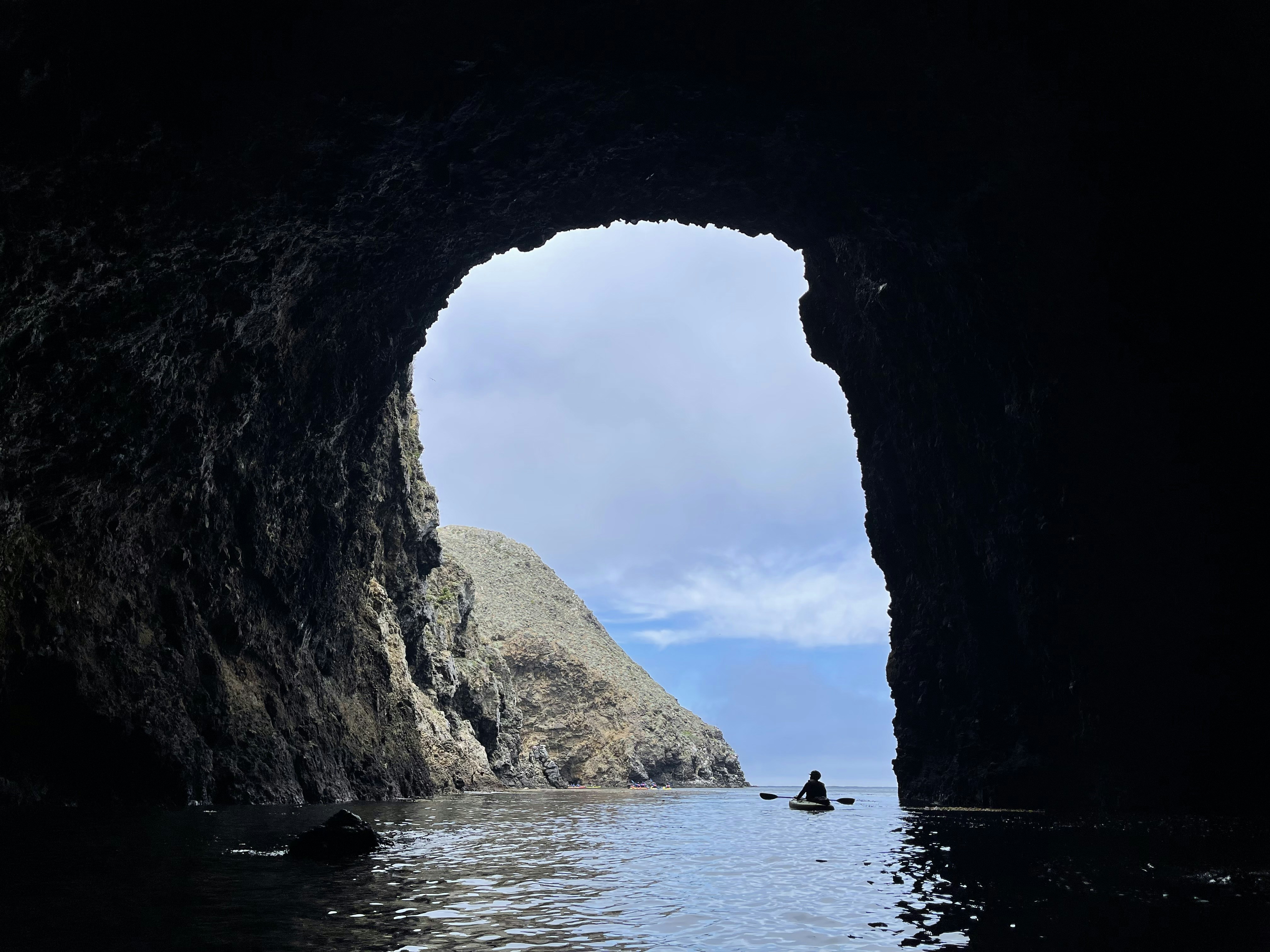 A lone kayaker paddles through a dramatic natural arch, framed by rugged cliffs and a serene sea, under a partially cloudy sky.