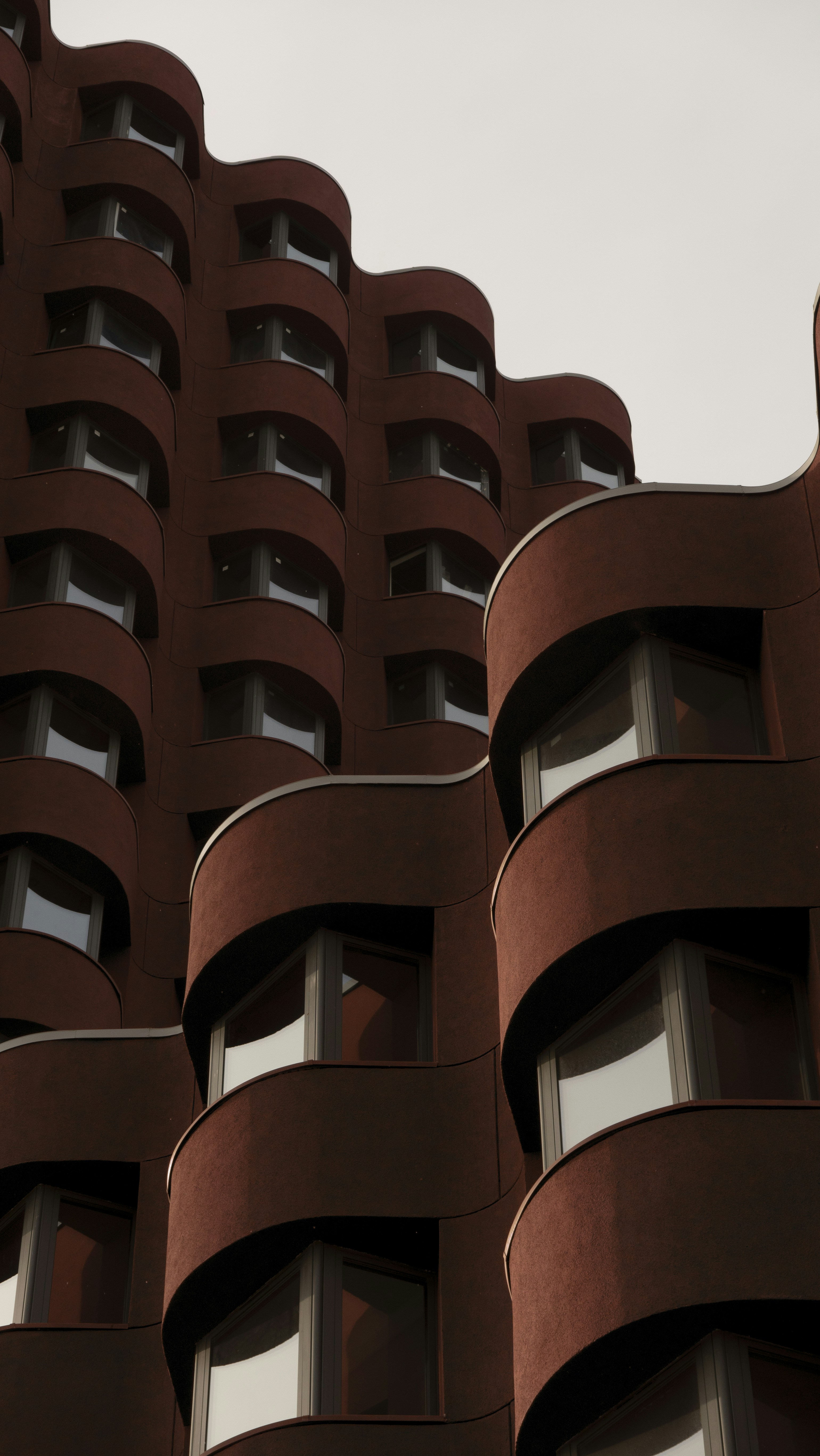 Brick-red façade features stacked rounded balconies forming a wave-like rhythm. The shot emphasizes geometric repetition and architectural texture.