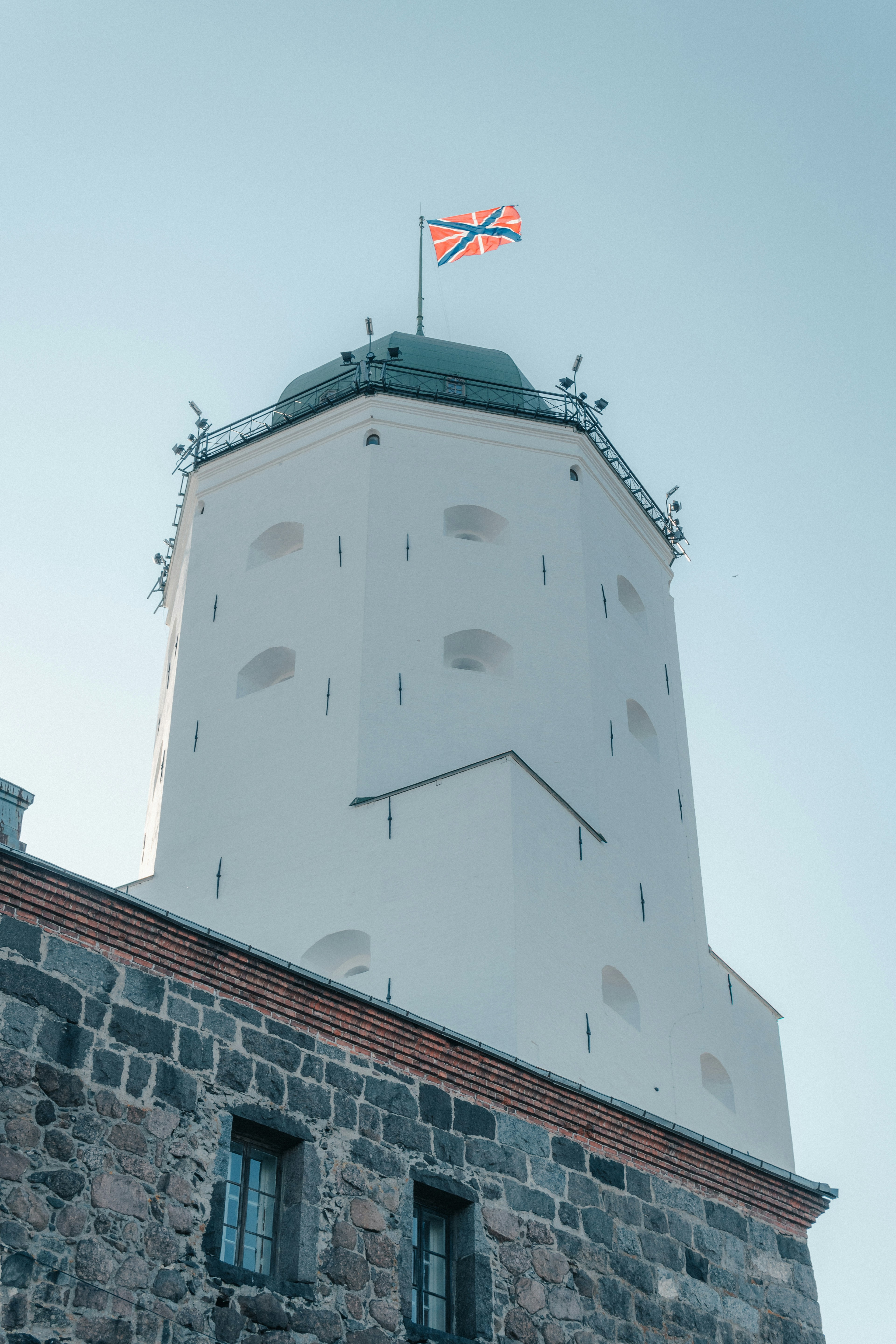 Flag atop a fortress tower: a white sentinel above rugged stone under a pale blue sky