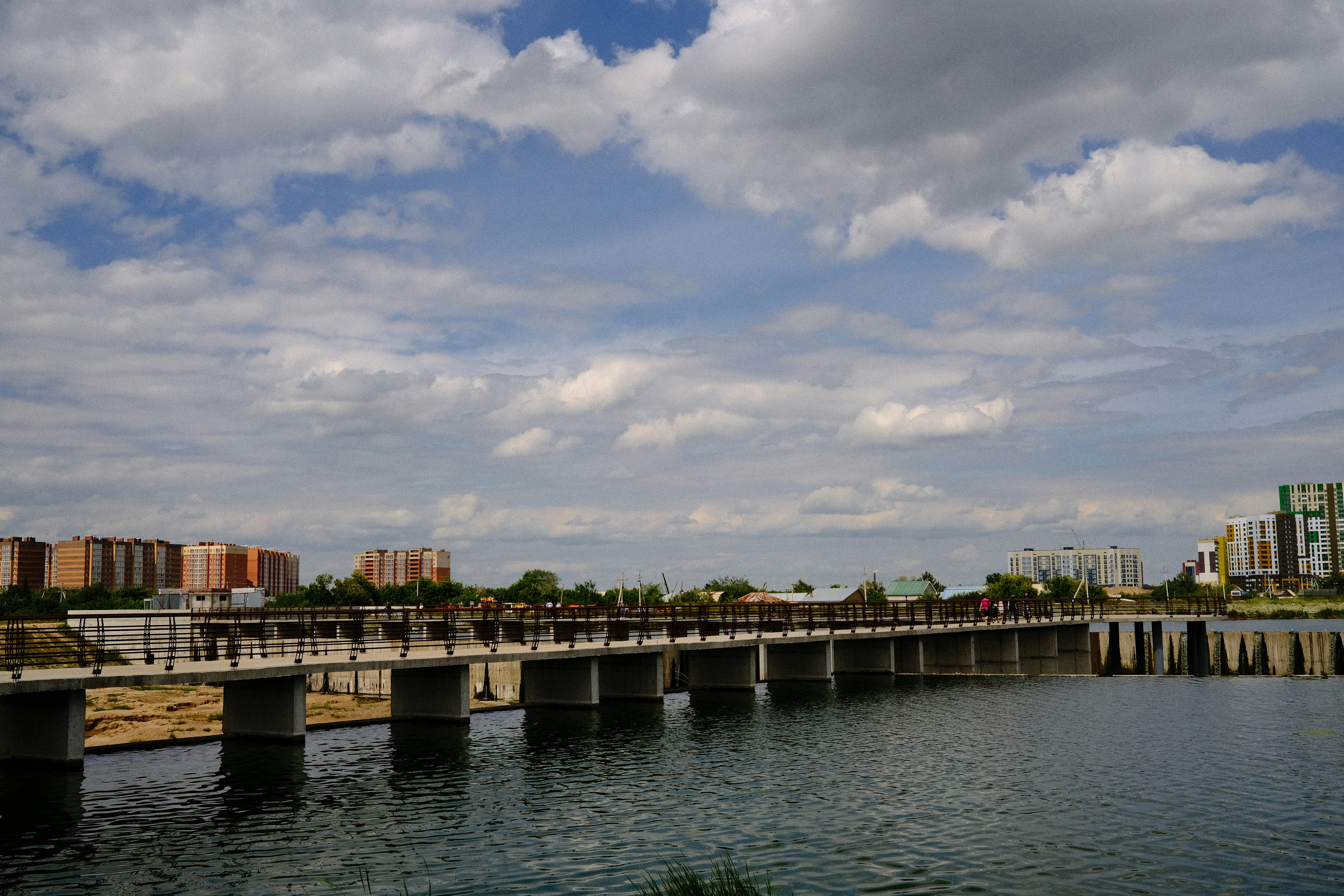 Concrete bridge spanning a tranquil river with urban buildings in the background under a partly cloudy sky.