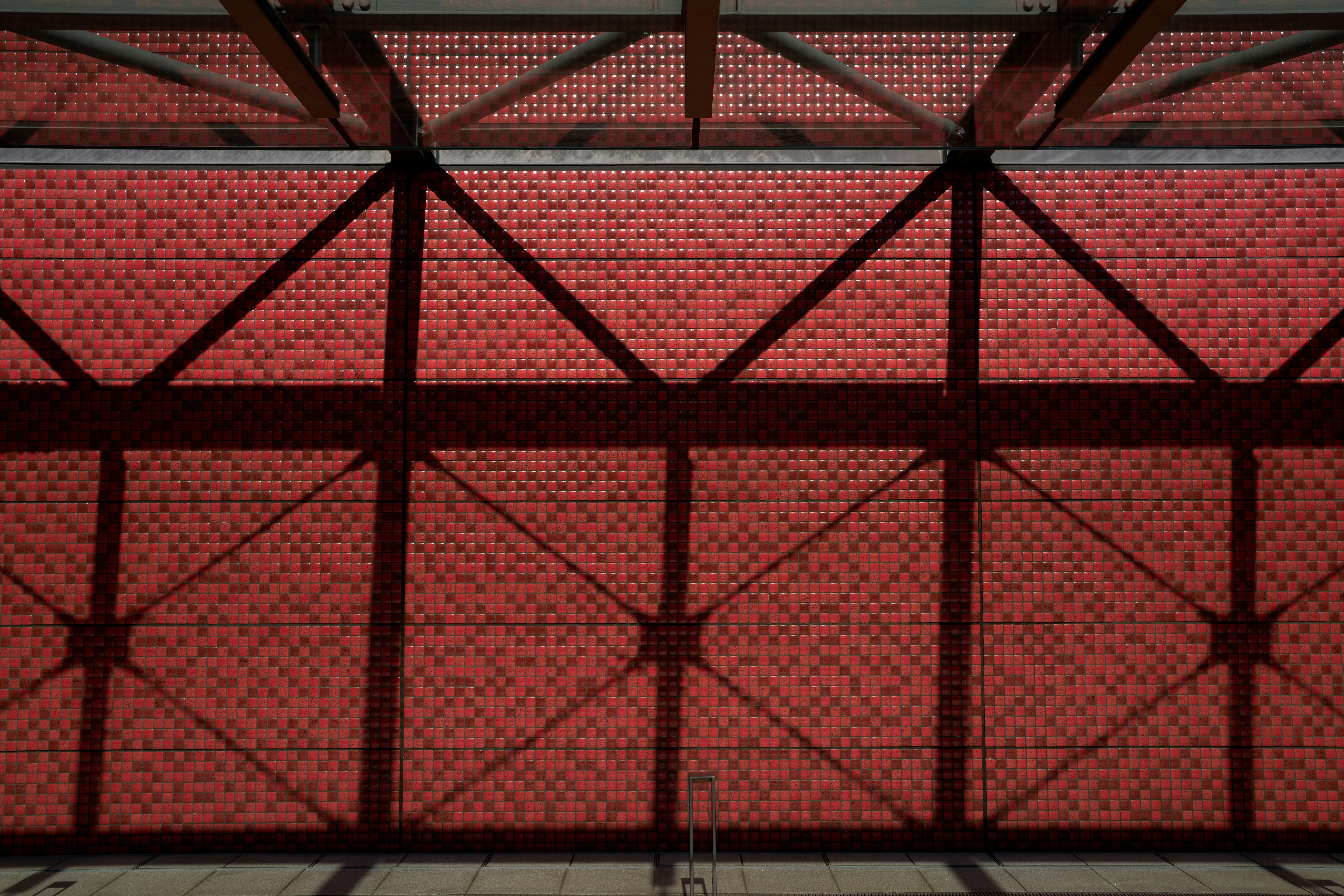 Intricate patterns of red tiles cast shadows through a modern architectural structure. The interplay of light and geometry creates a visually striking composition.