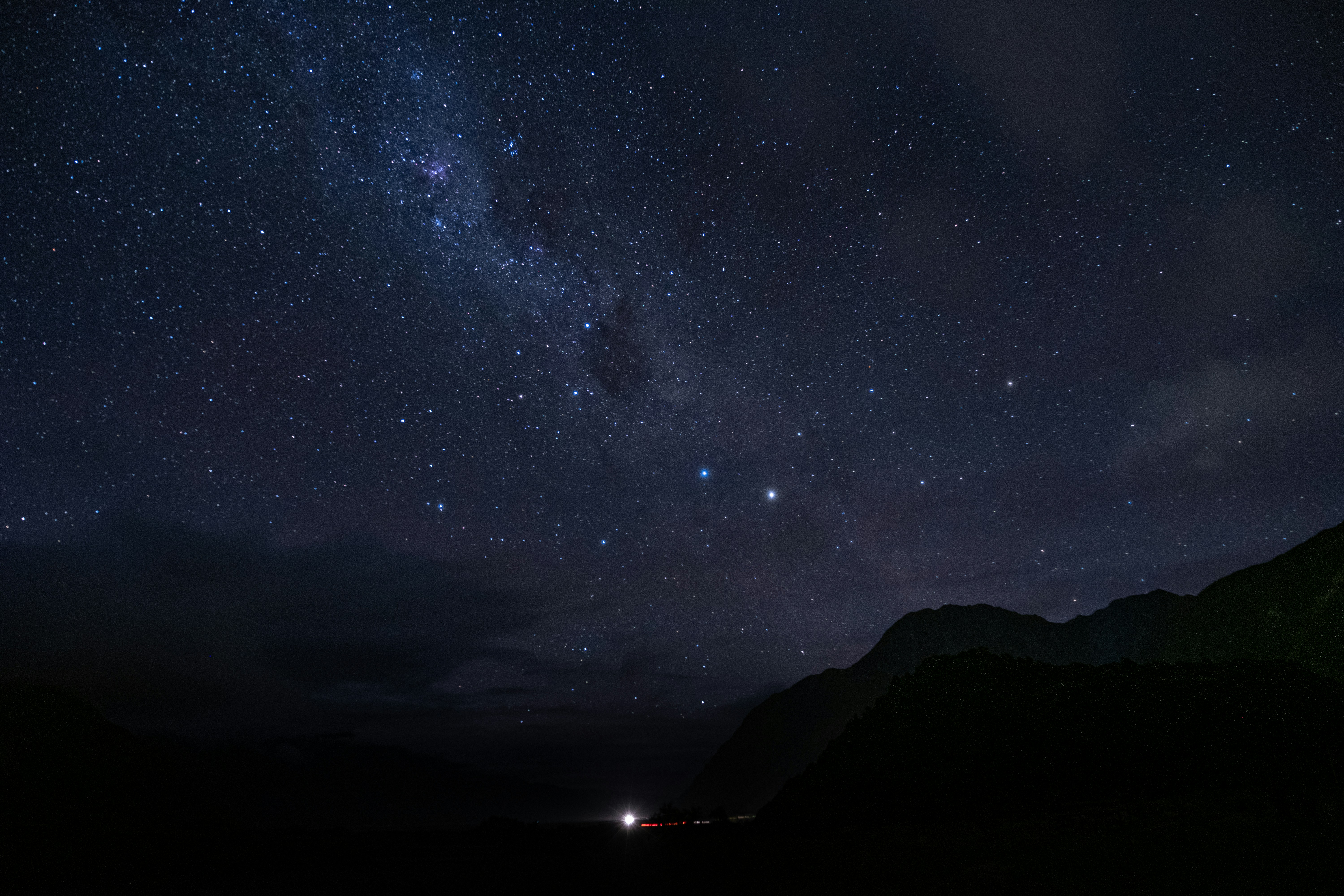 Star-filled sky arches over dark mountain silhouettes with faint lights on the horizon.