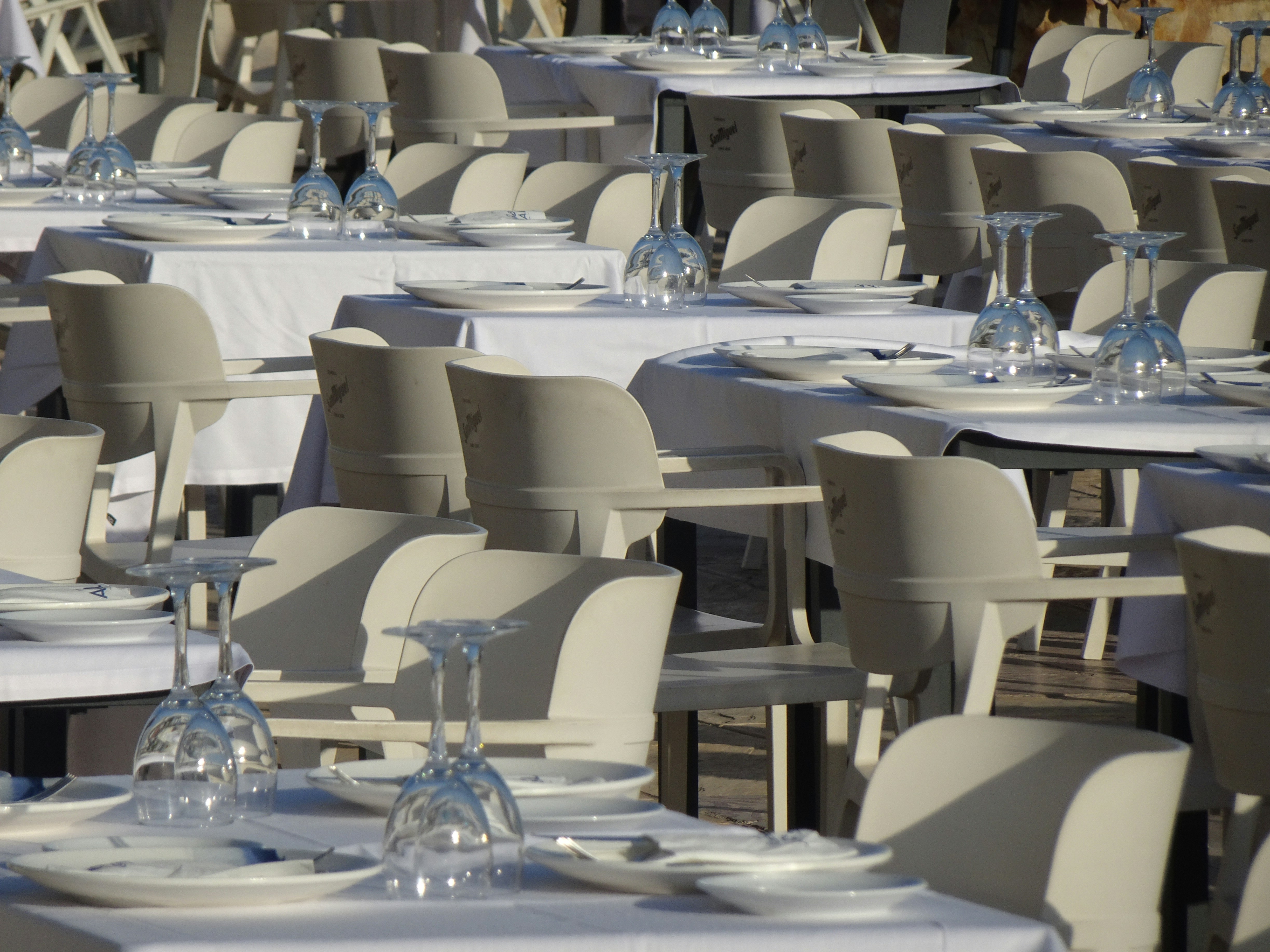 Sunlit outdoor dining area with neatly arranged white tables, chairs, and inverted glassware set for service.