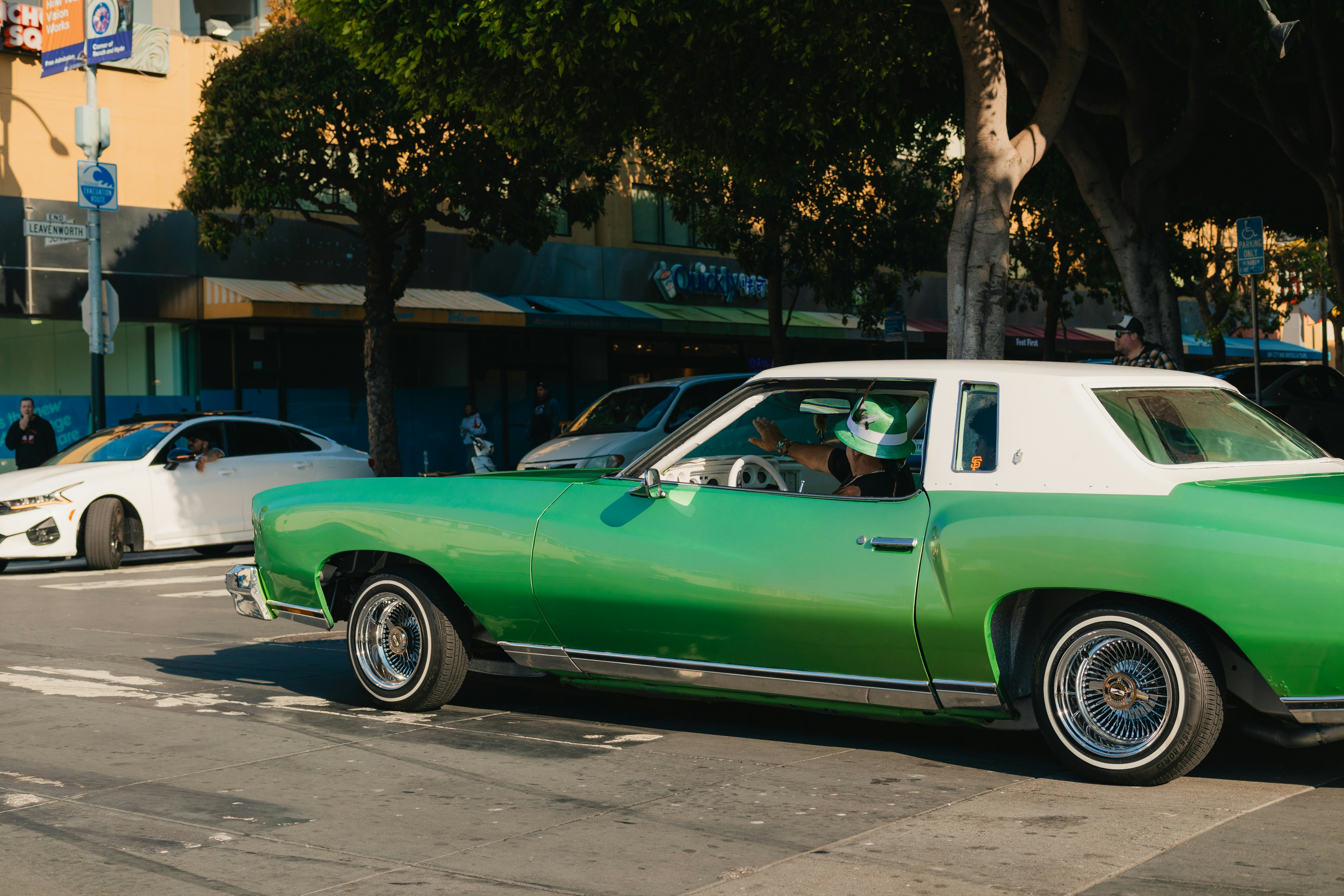 Green vintage car cruising past a sunny storefront row