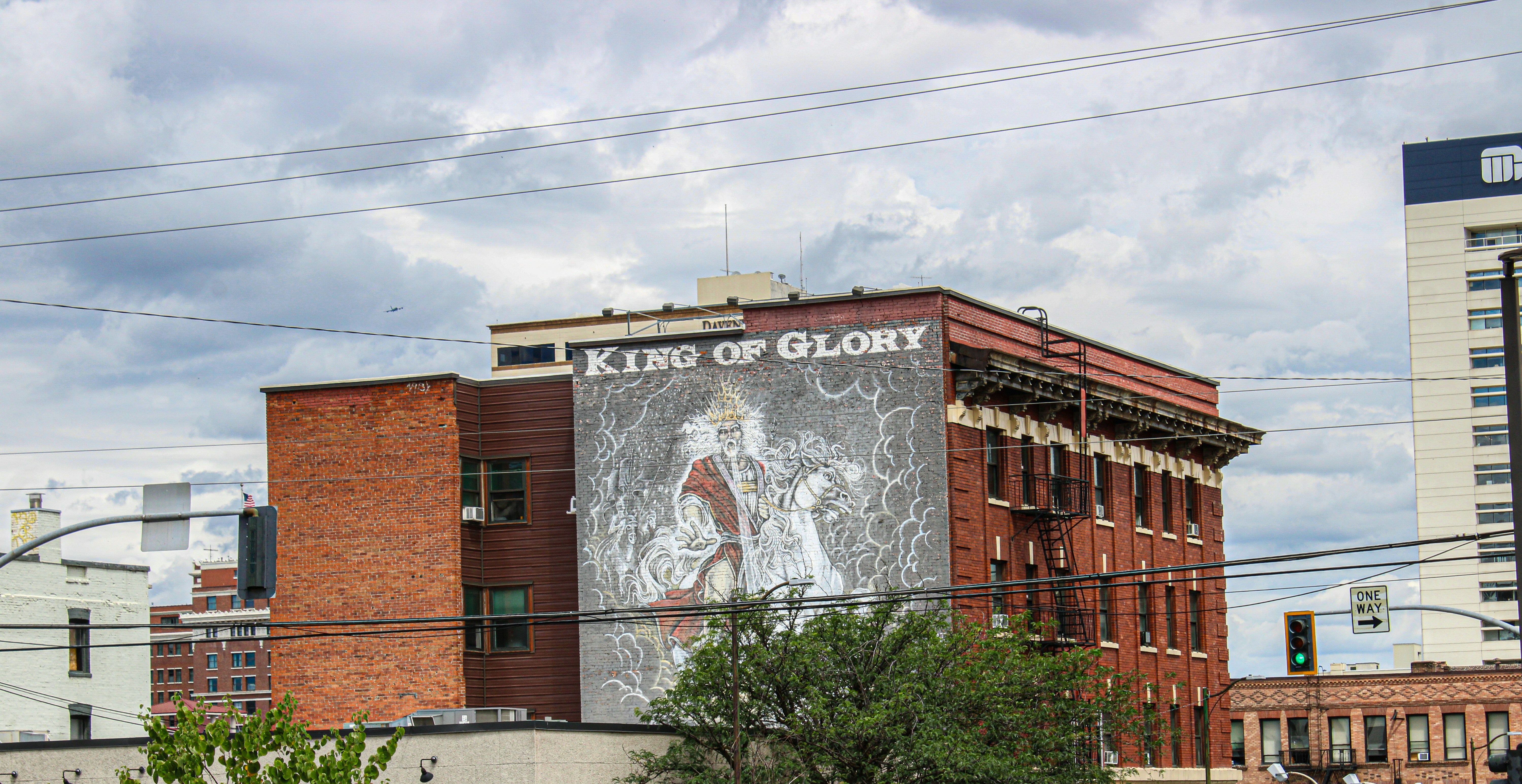 Street mural depicting a regal figure on a horse, titled 'King of Glory,' on a brick building under a cloudy sky.