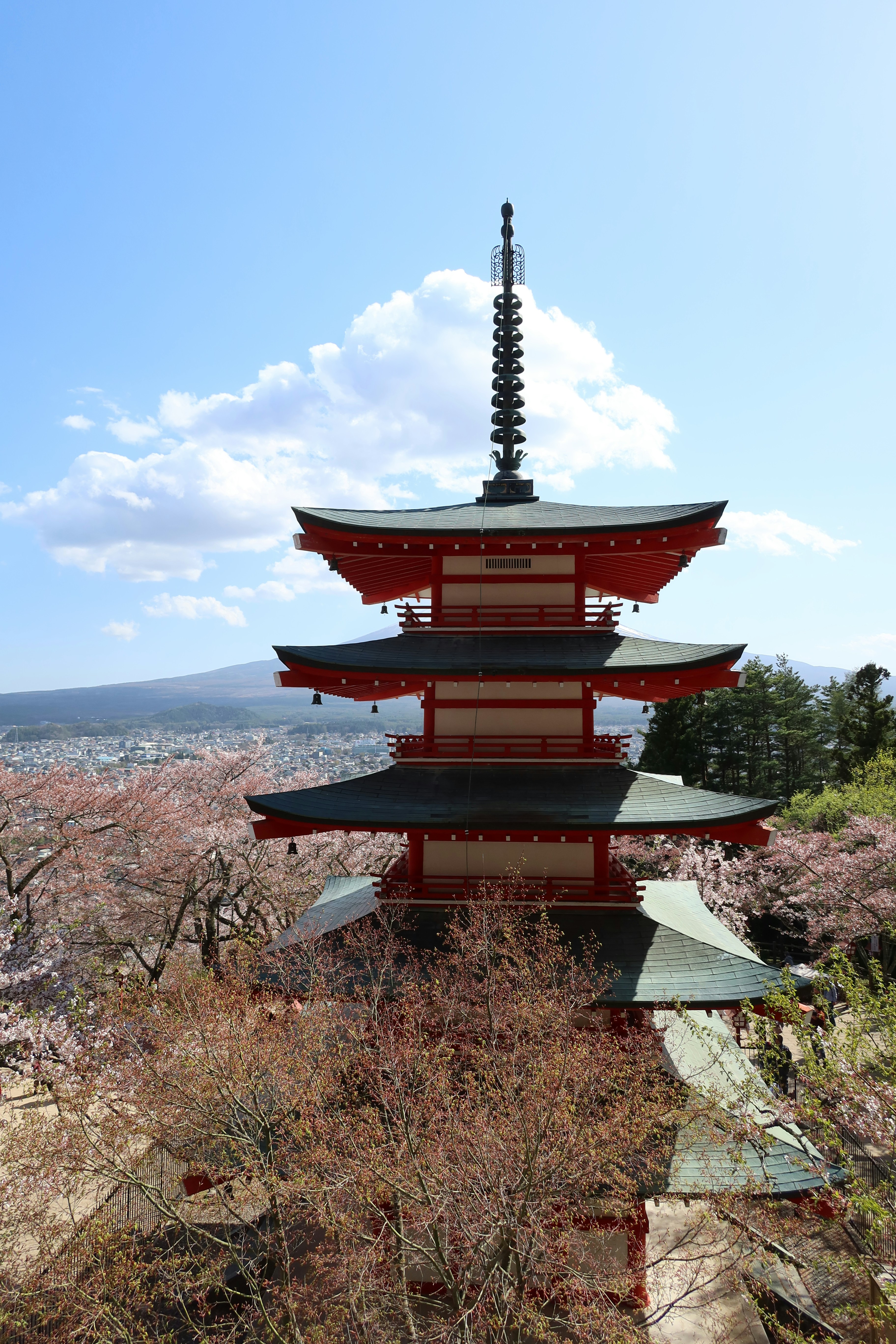 Five-story red pagoda rises above blooming cherry blossoms against a clear blue sky. Distant hills and a town spread along the horizon.