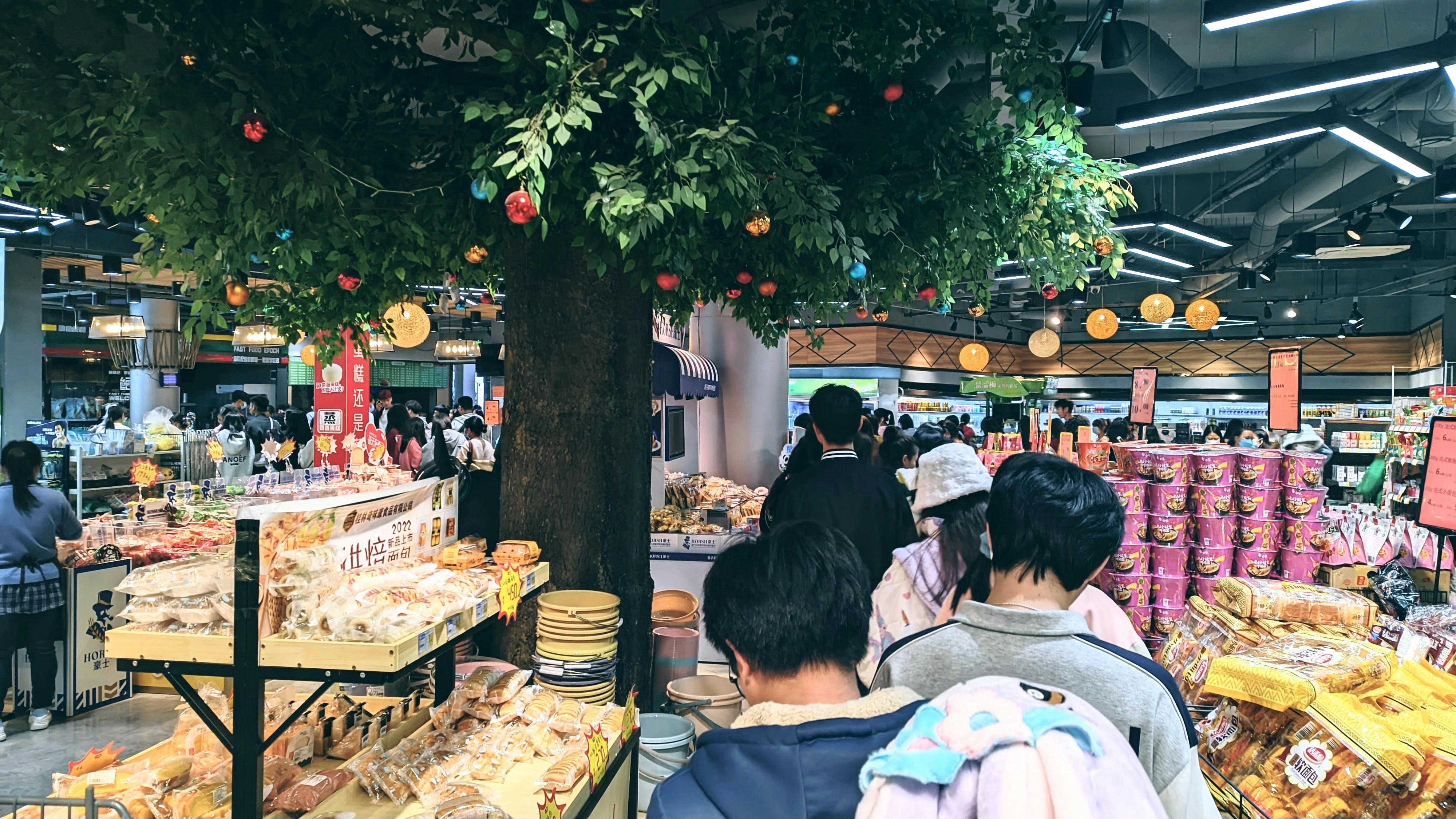 Vibrant market scene featuring shoppers navigating through aisles filled with colorful products and a decorative tree overhead.