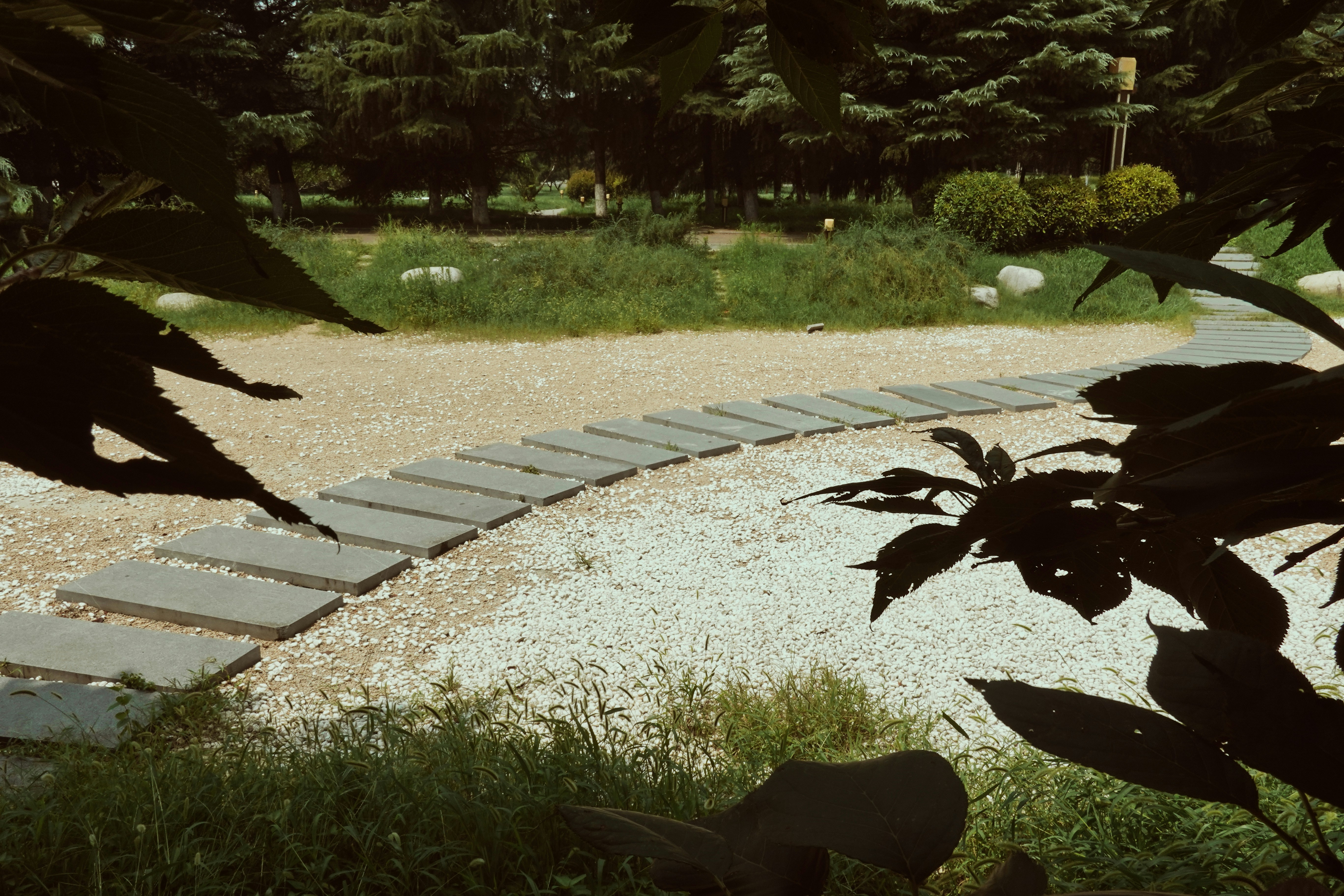 A path in a park, the pavement is made of neatly arranged stone slabs, surrounded by thick trees and grass. The path curves forward, and the plants on both sides form a natural arch, giving a quiet, peaceful feeling. This design is both beautiful and practical, allowing people to enjoy nature while walking easily.