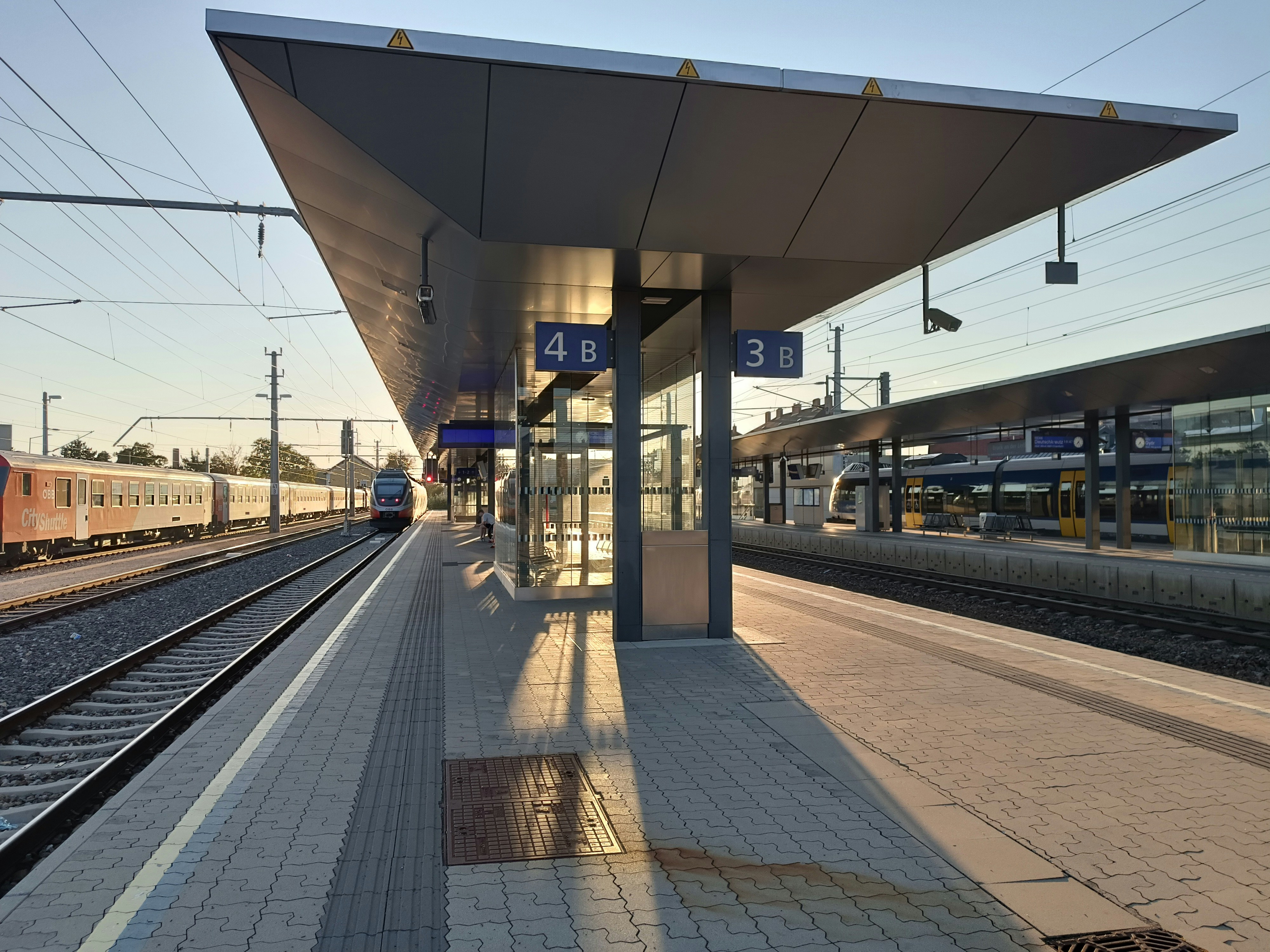 Train station platform with numbered signs, illuminated by the warm glow of sunset. Two trains are visible on adjacent tracks.