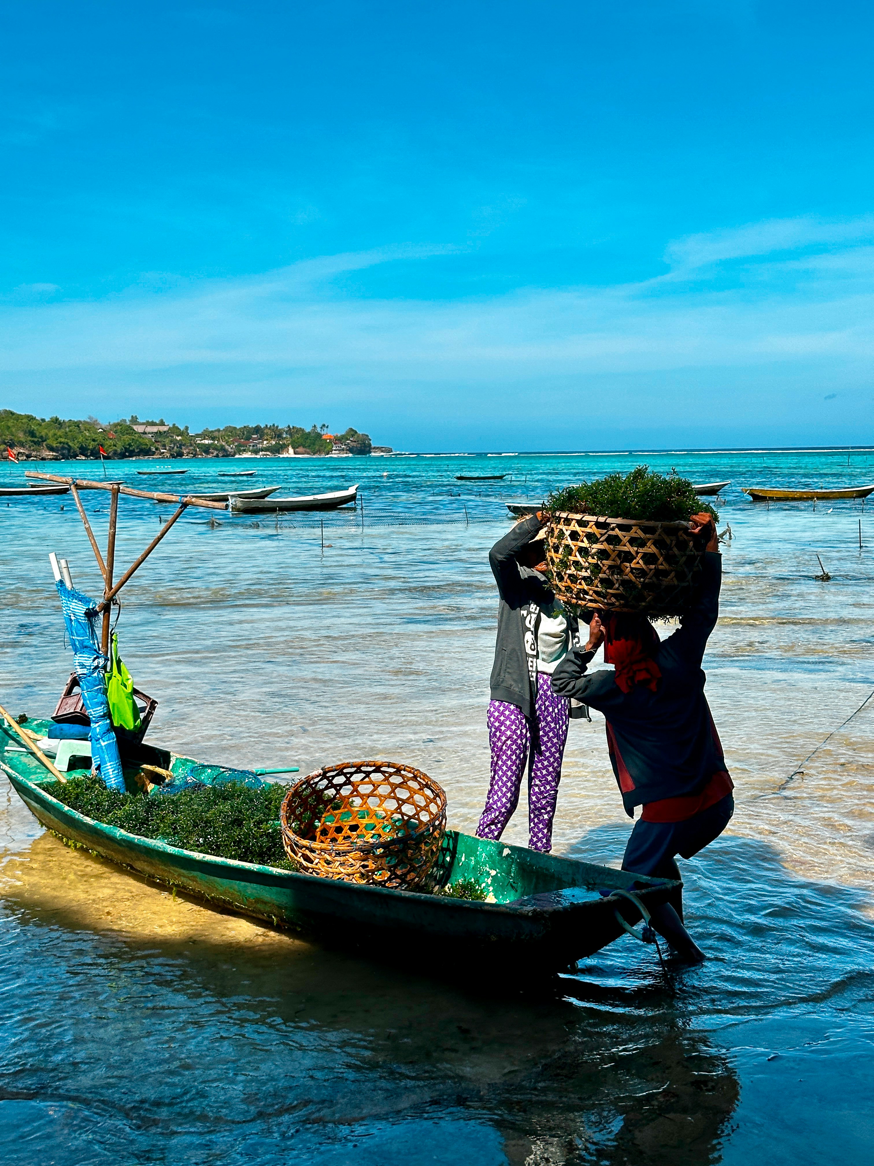 A woman is helping a man place a basket full of seaweed on his head as they stand by a small boat in the afternoon sun.