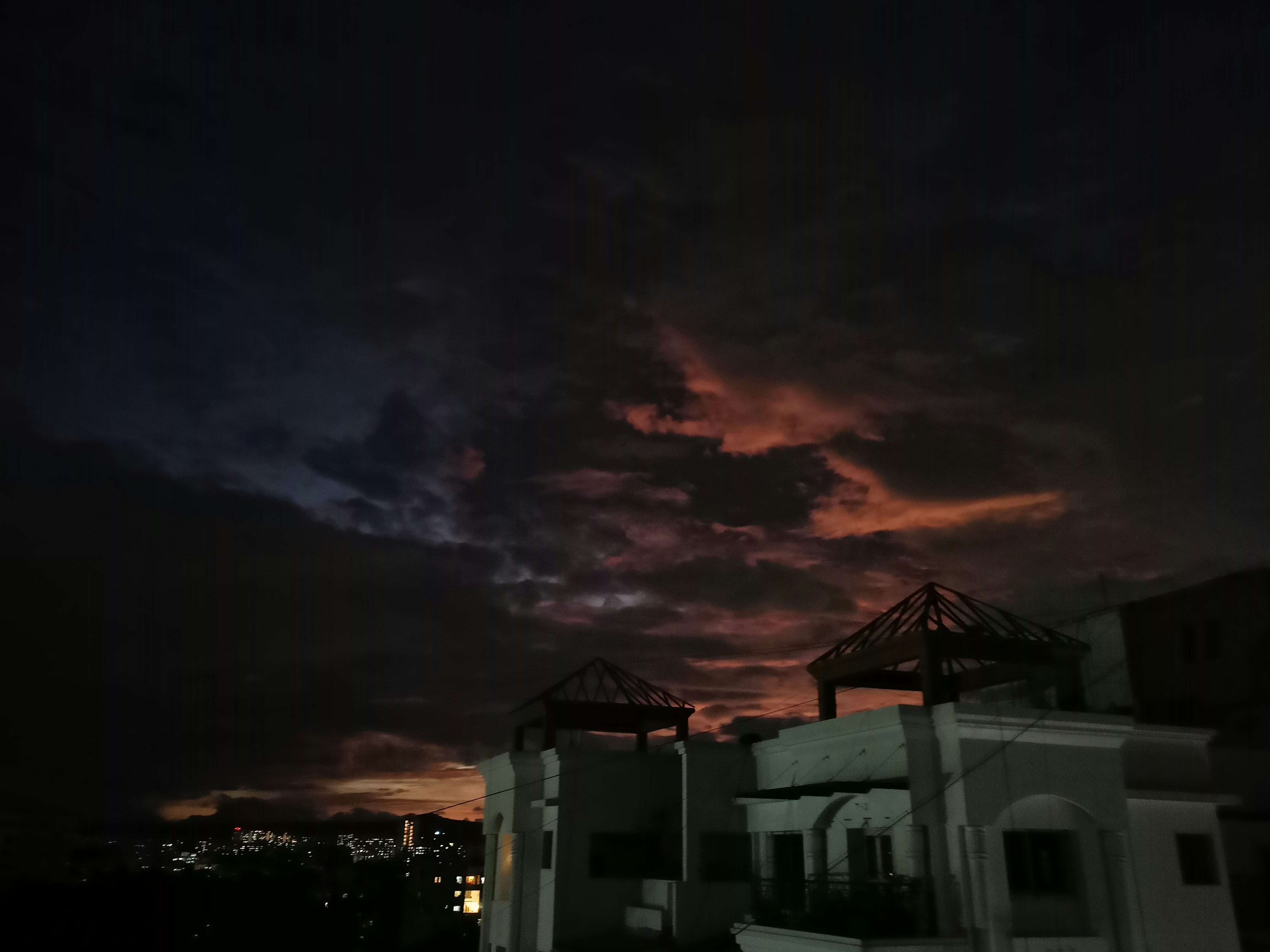 Dark rooftop silhouettes beneath a dusk sky lit by red-orange clouds and distant city lights.