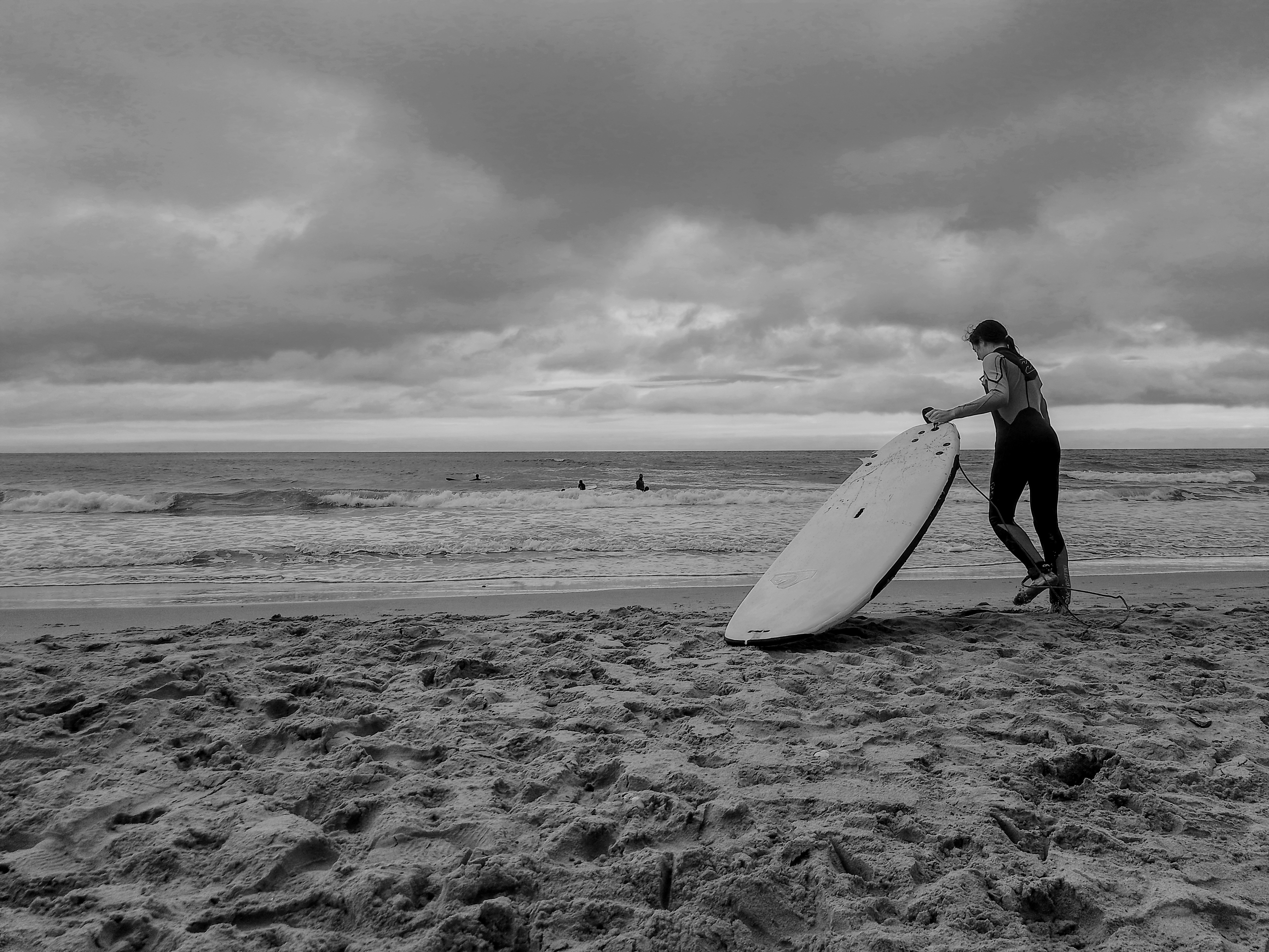 Surfing at Seafield Bay, Tara Cove, Wexford, Ireland. A black-and-white photograph capturing a lone surfer on a sandy beach as they prepare to enter the ocean. The surfer, clad in a wetsuit, drags their board against the backdrop of a moody, cloud-filled sky and gentle waves rolling onto the shore. The textured sand in the foreground and the vast, open sea in the background evoke a sense of solitude and connection with nature, emphasizing the raw beauty of coastal life.
