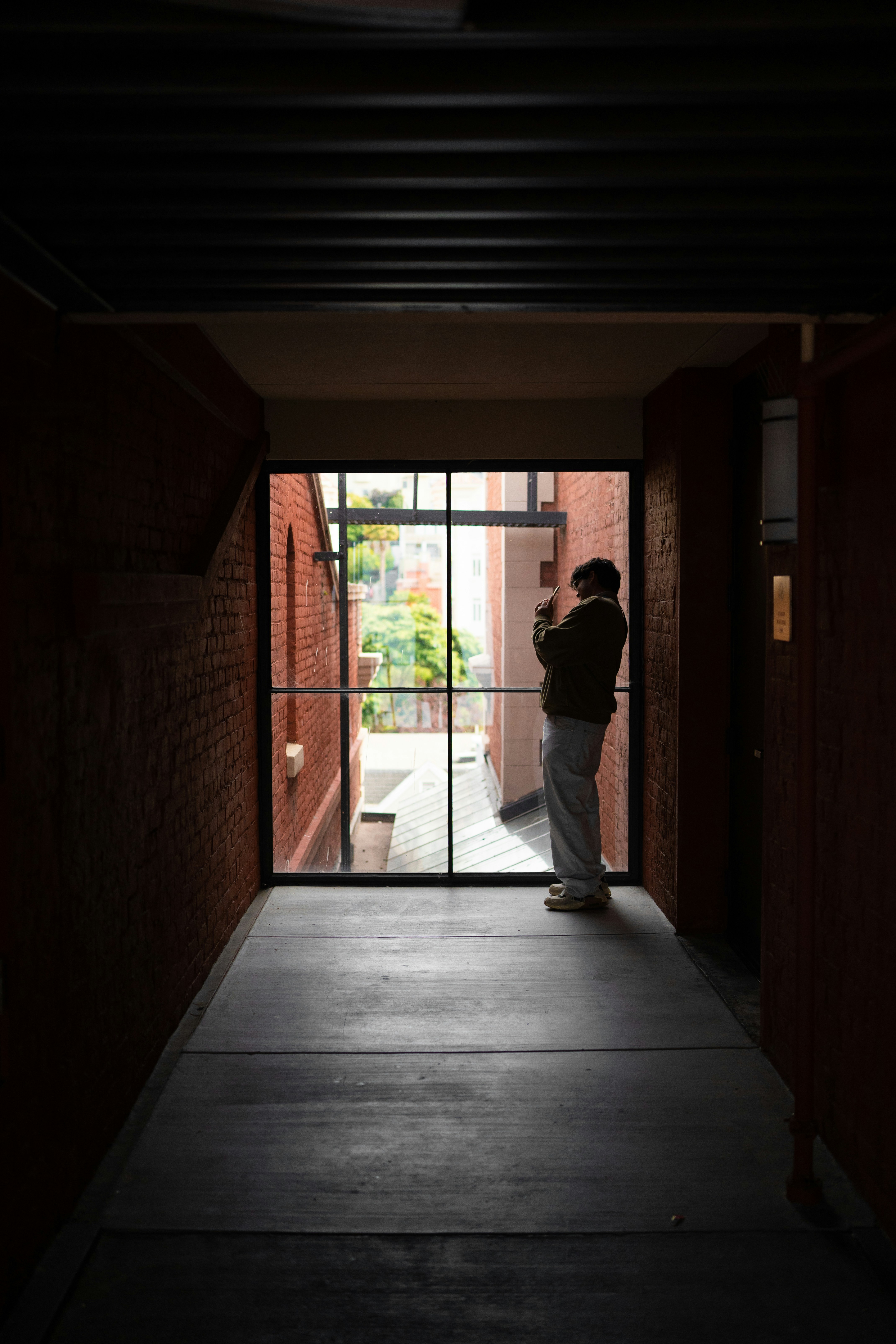 Urban Threshold: A solitary photographer framed by brick walls as daylight spills through a glass window