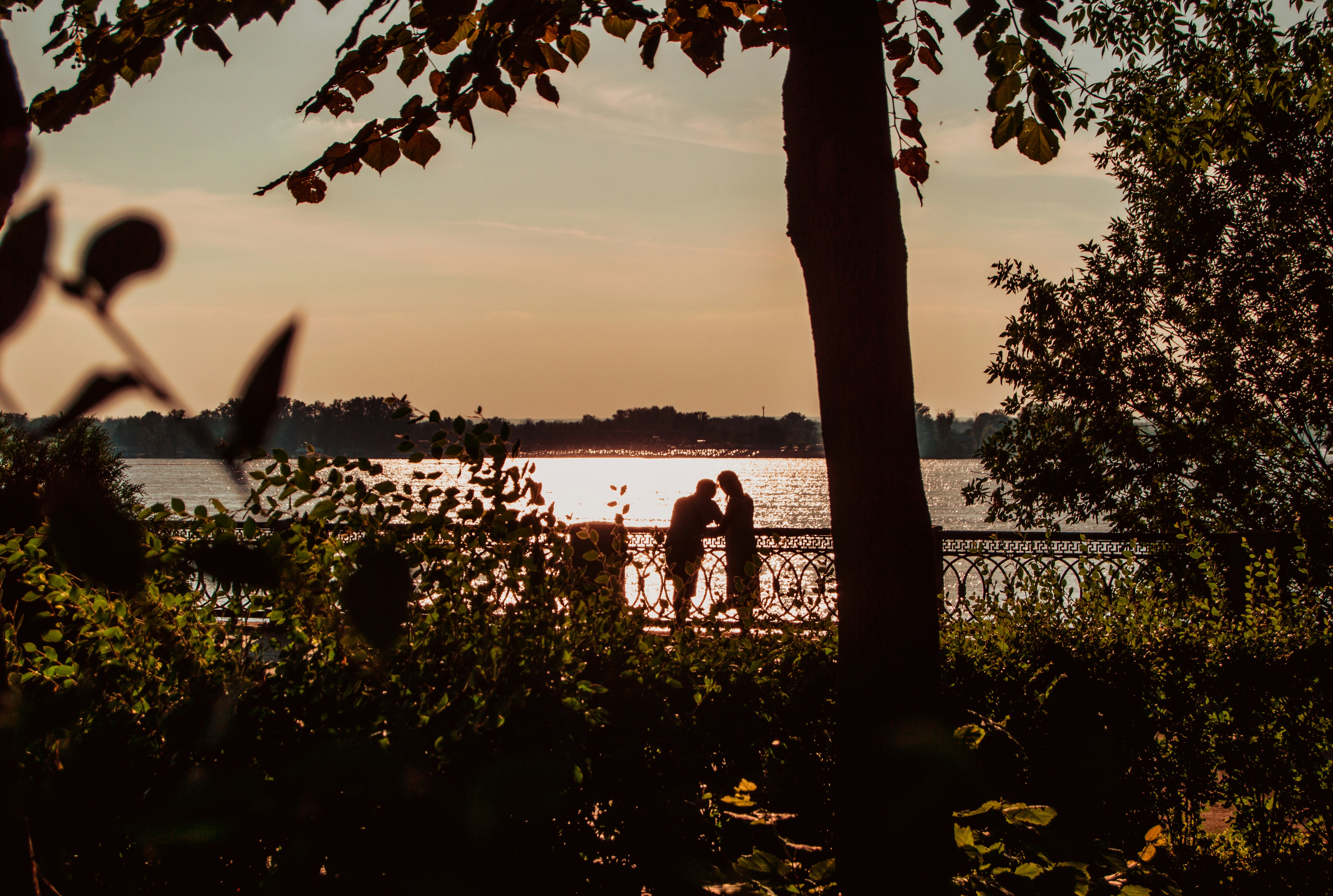 Silhouetted couple stands by a lakeside, surrounded by trees, as the sun sets on the horizon.