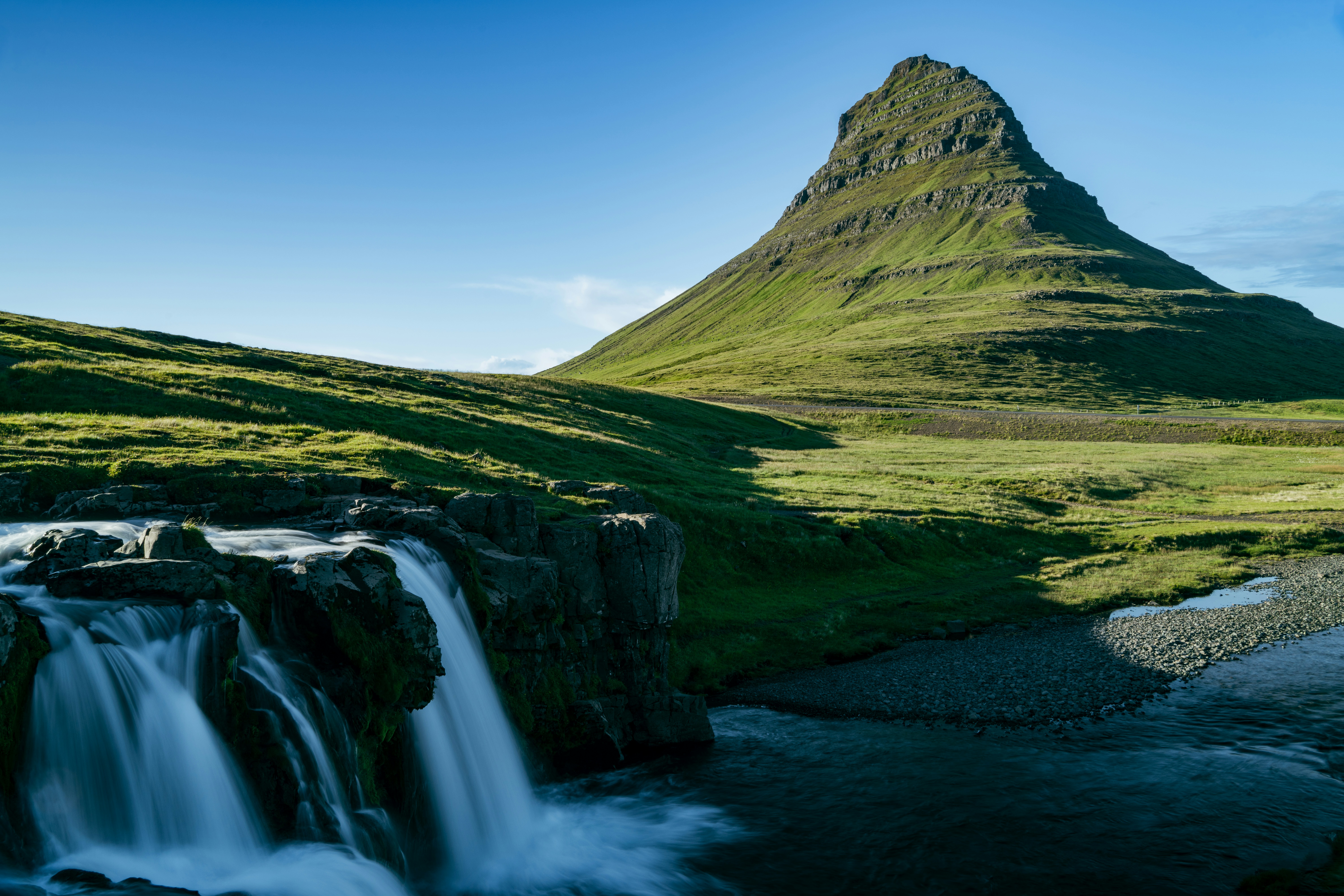 Kirkjufell mountain rises sharply against a clear sky, with a gentle waterfall flowing over rocks in the foreground.