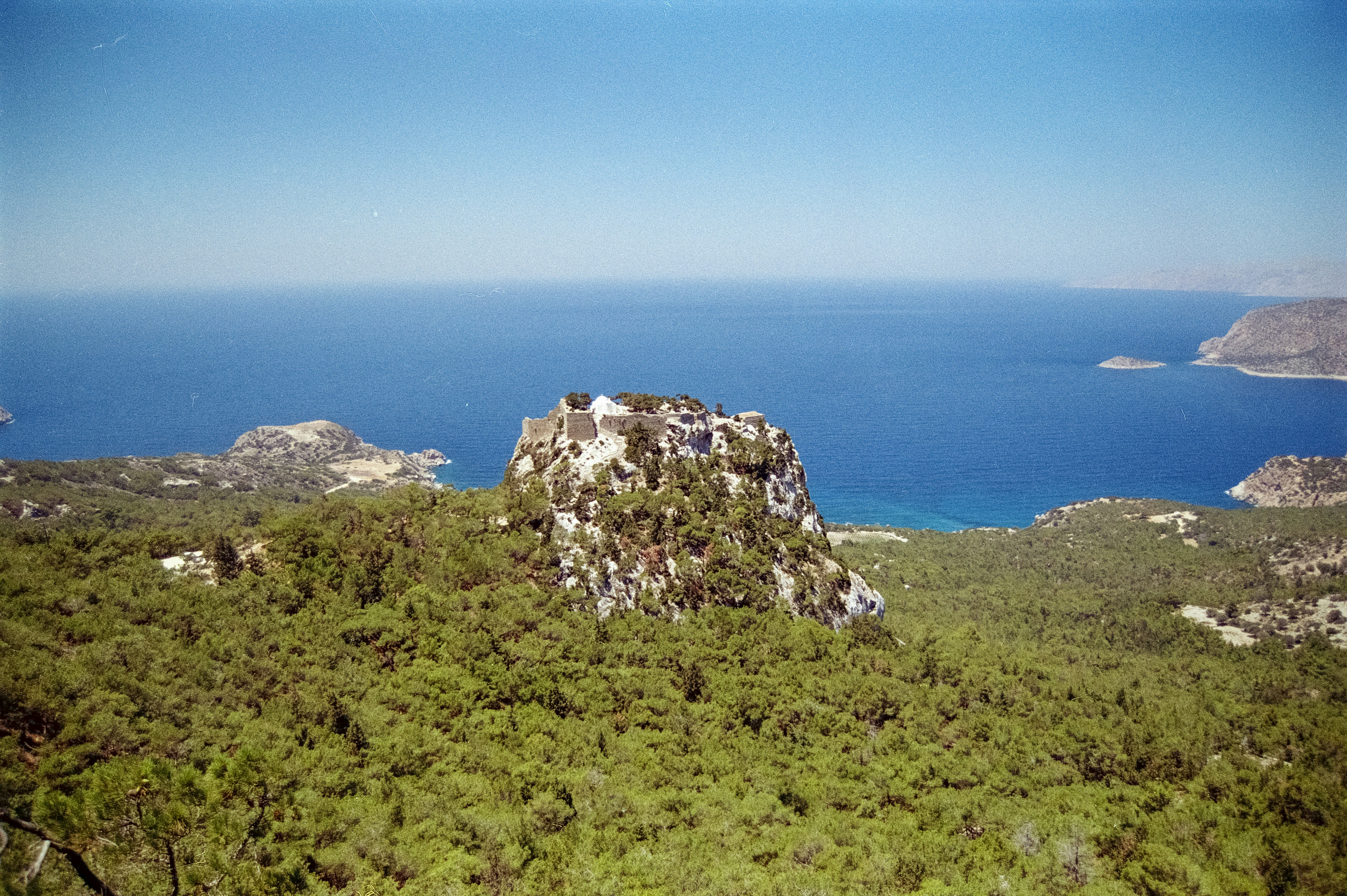 Hilltop fortress surrounded by lush green forest with a vast blue sea in the background.