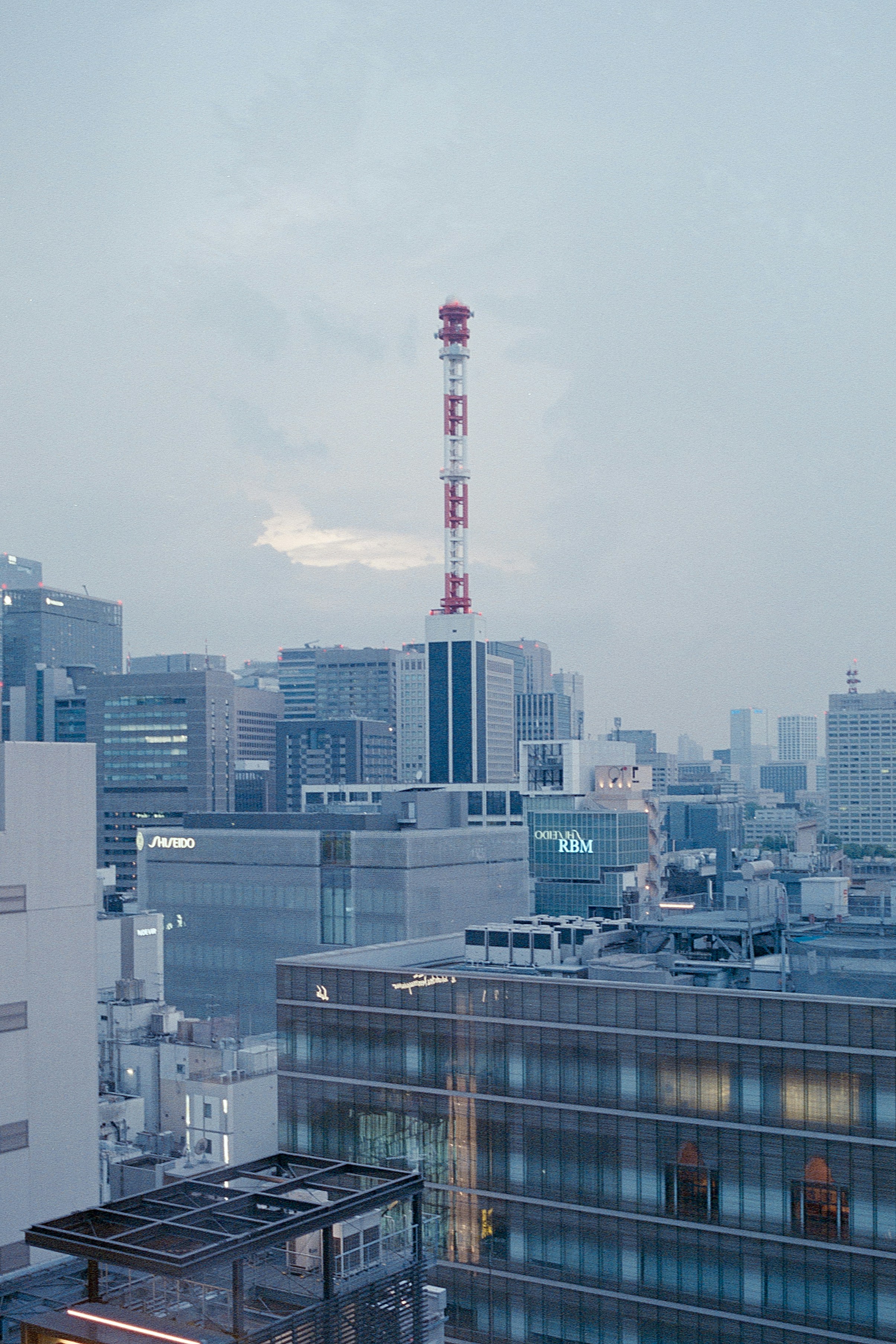 Cityscape photograph featuring a tall red-and-white antenna rising above glass-fronted office buildings at dusk, with a hazy sky behind.