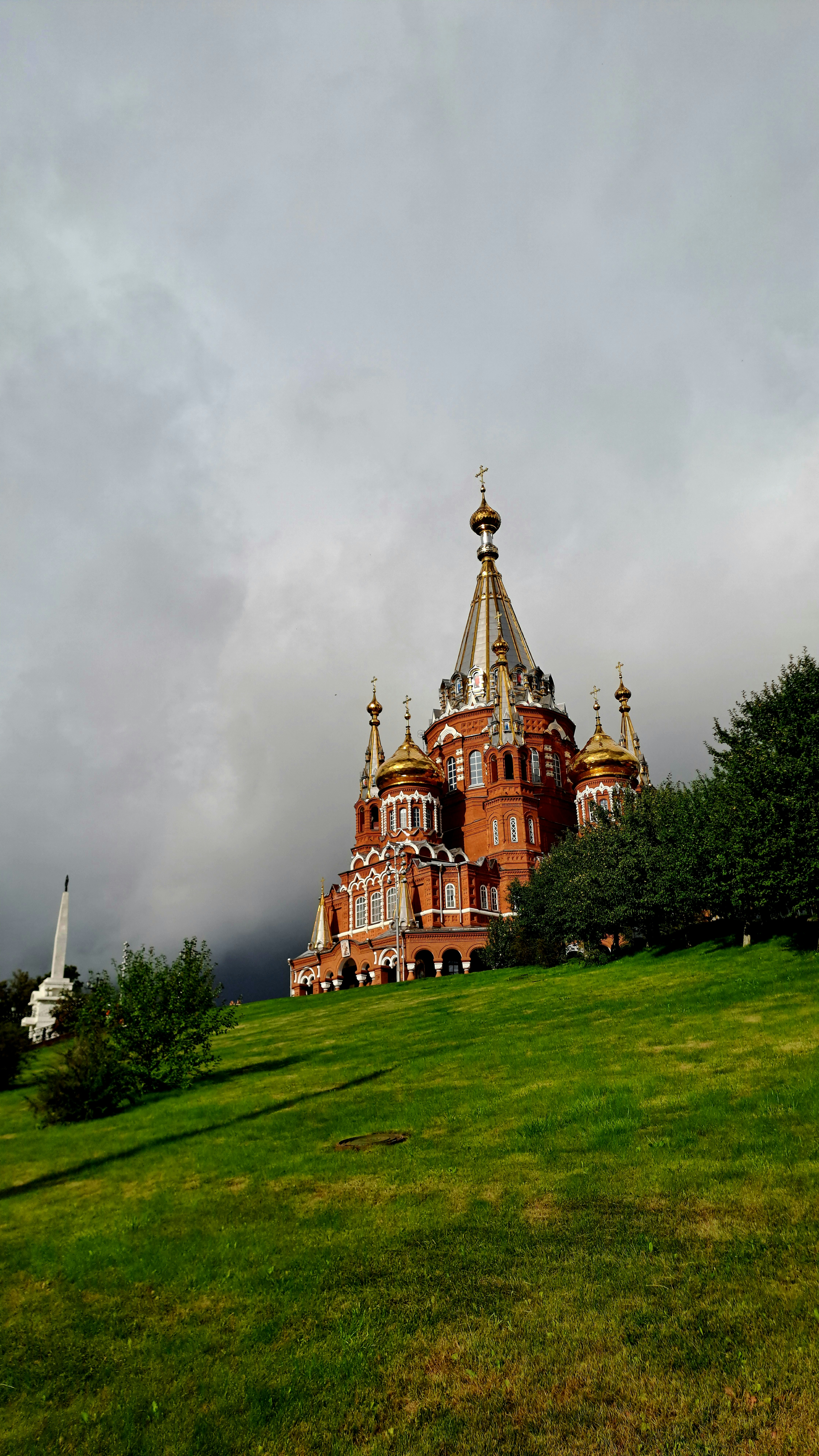Intricate red-brick church with golden domes rises against a dramatic, cloudy sky. Lush green grass frames the scene, hinting at a serene yet dynamic atmosphere.