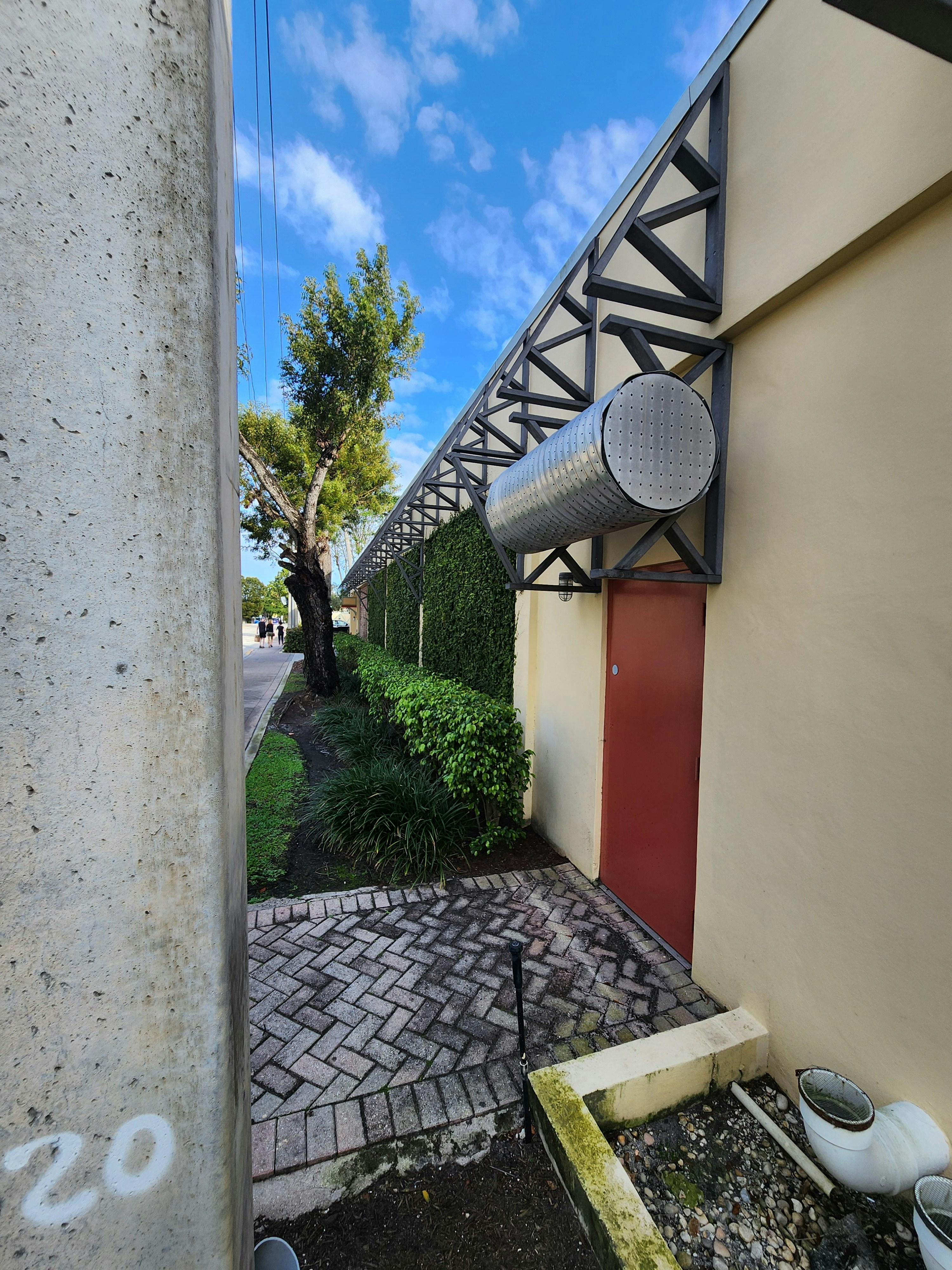Ductwork Framing a Sunlit Corridor: Door, Hedge, and Brick Path