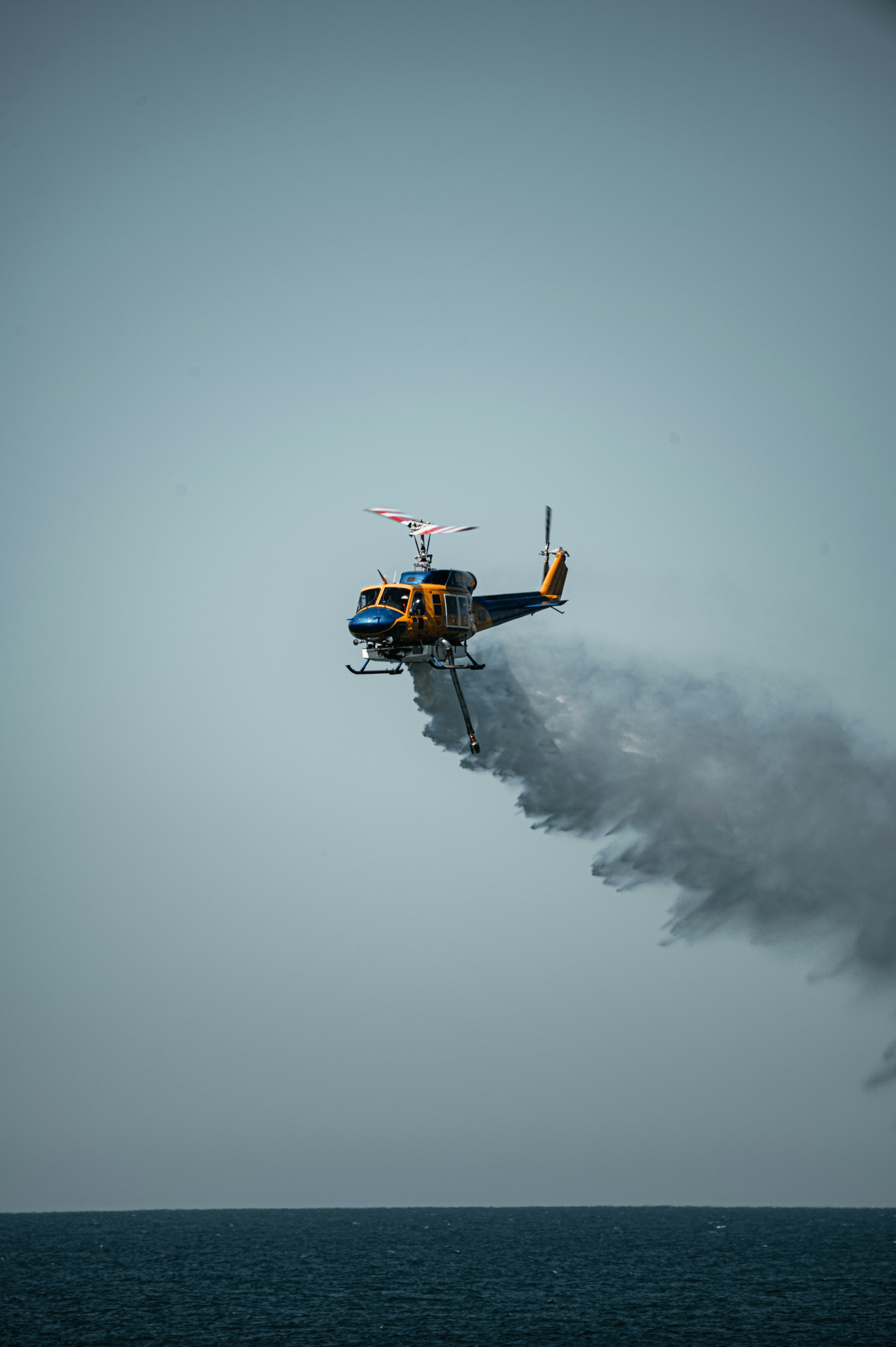 Coastal Blaze Aerial: Yellow firefighting helicopter threads a smoke trail over the sea