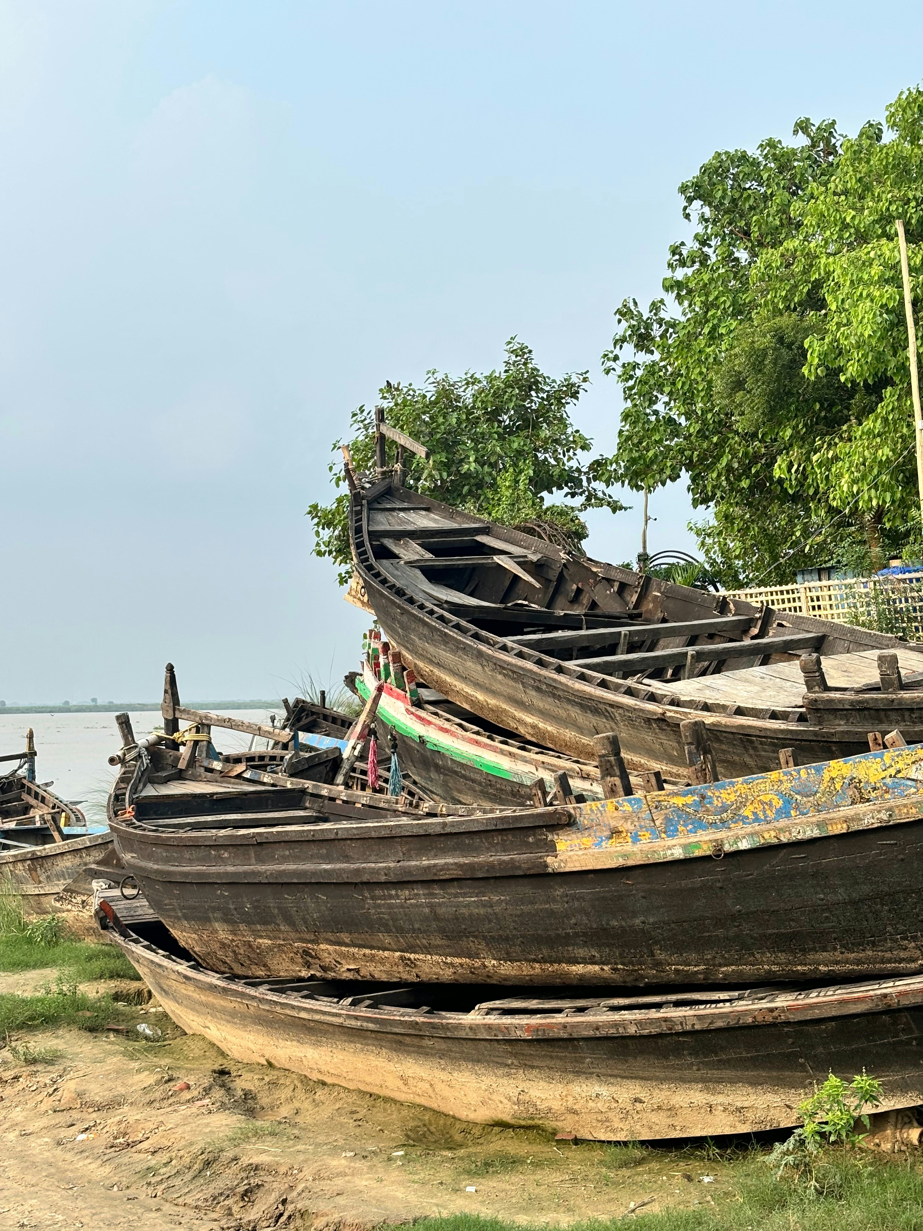 A cluster of old wooden boats stacked on a sandy shore beside a calm lake, with green trees in the background.