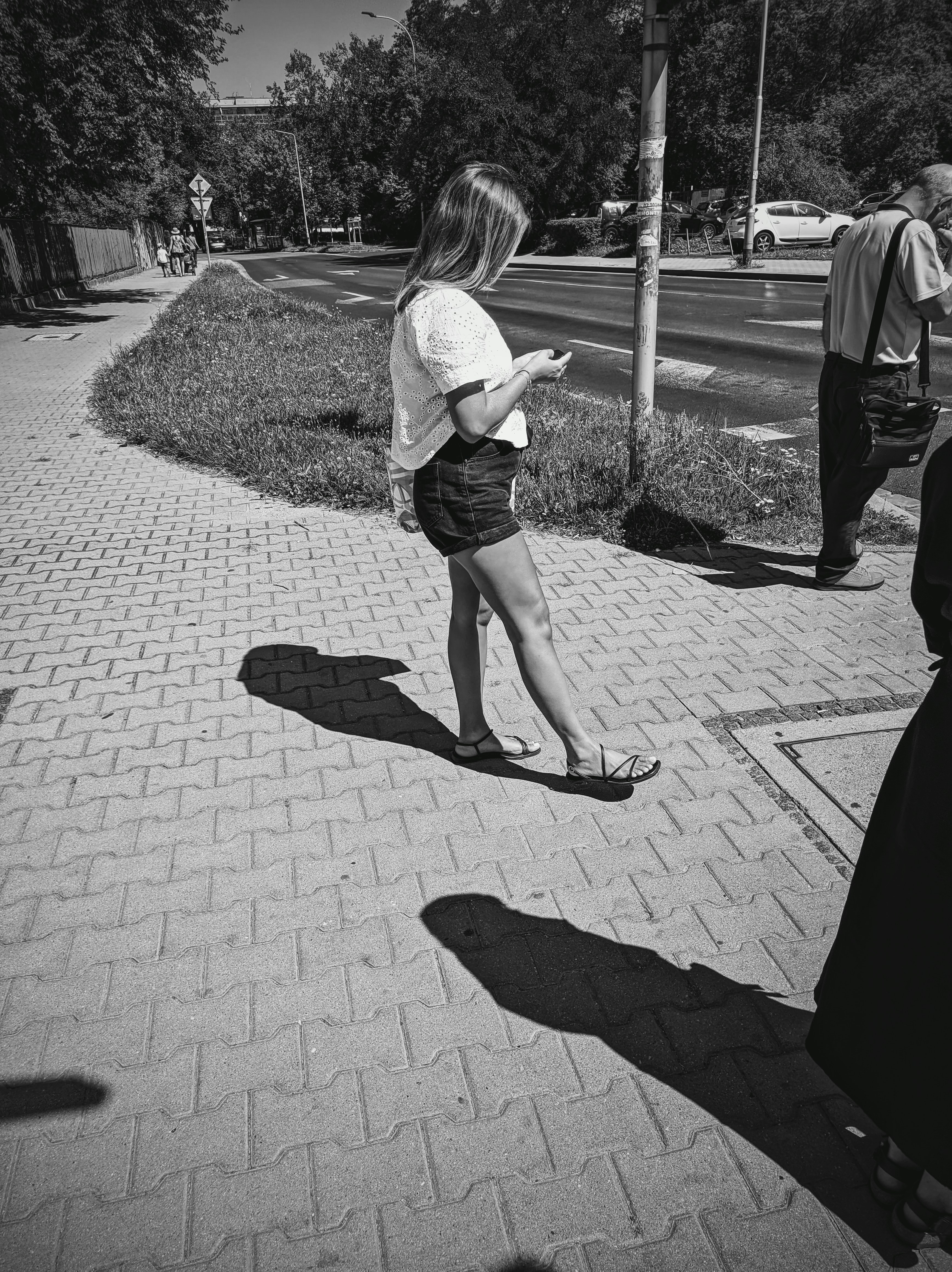 Photograph of a woman in a white top and shorts walking along a patterned sidewalk while checking her phone; bold shadows and an urban background define a candid street moment.