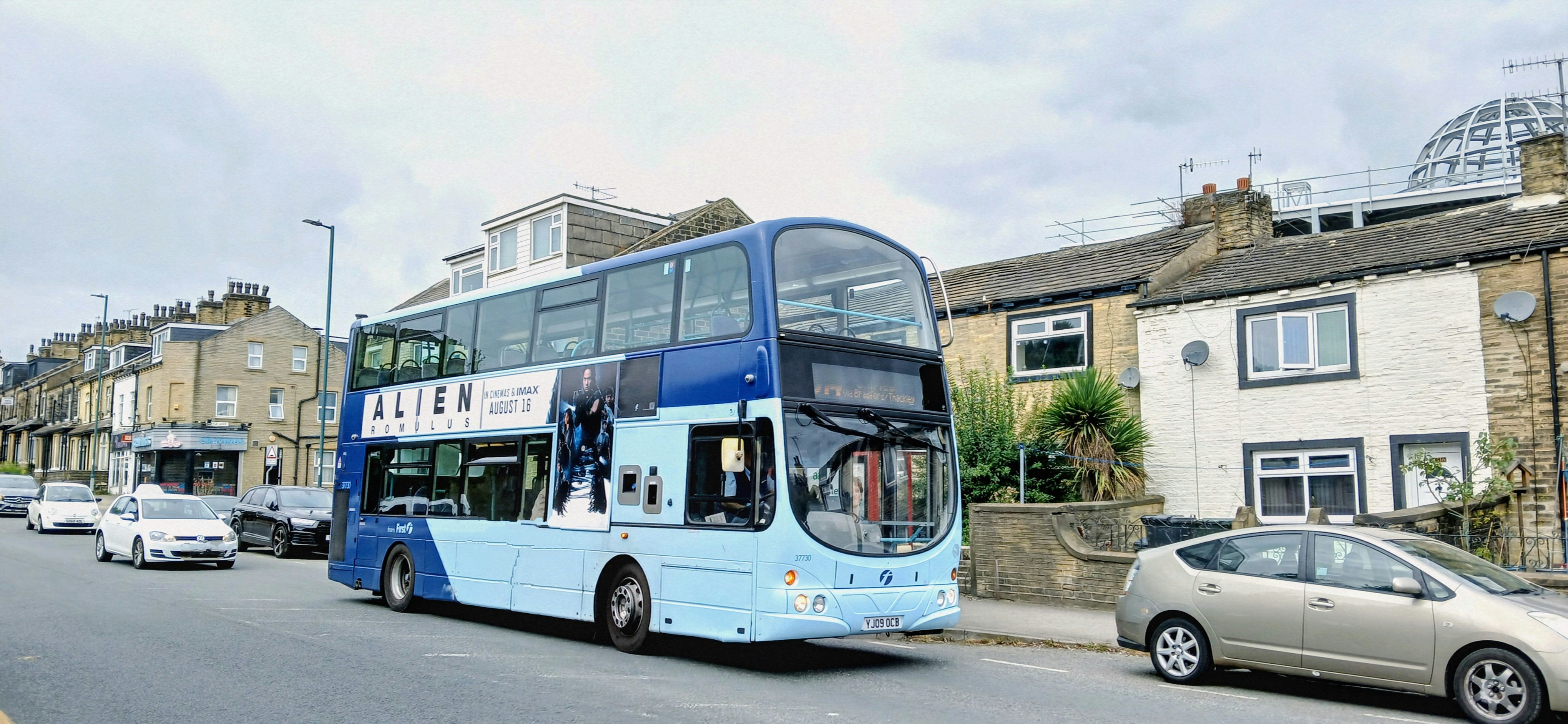 Blue double-decker bus glides past a row of pastel houses on a sunlit street.