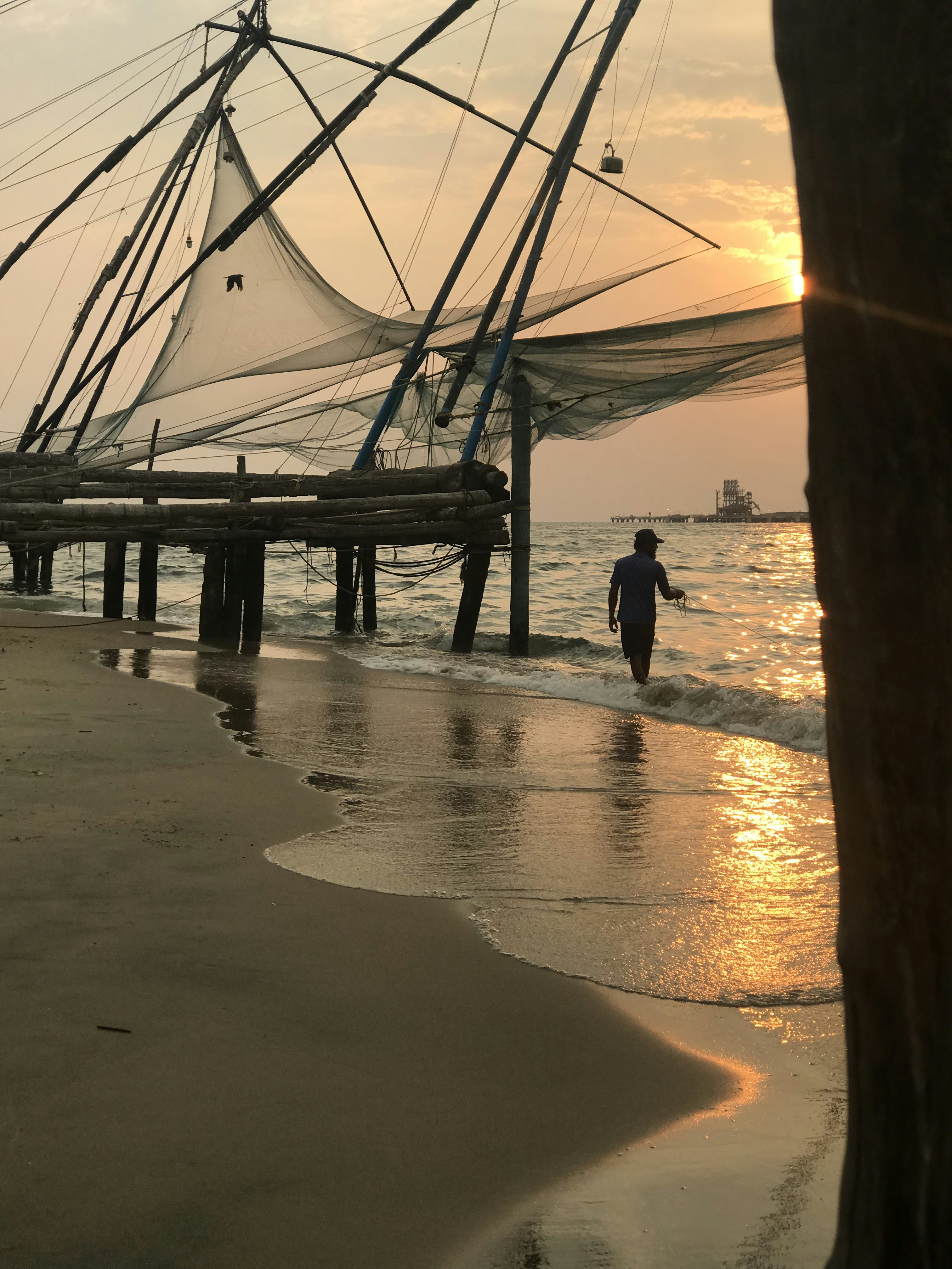 A lone figure walks along a sandy beach, framed by traditional fishing nets silhouetted against a glowing sunset. The rhythmic waves reflect the warm hues of dusk.