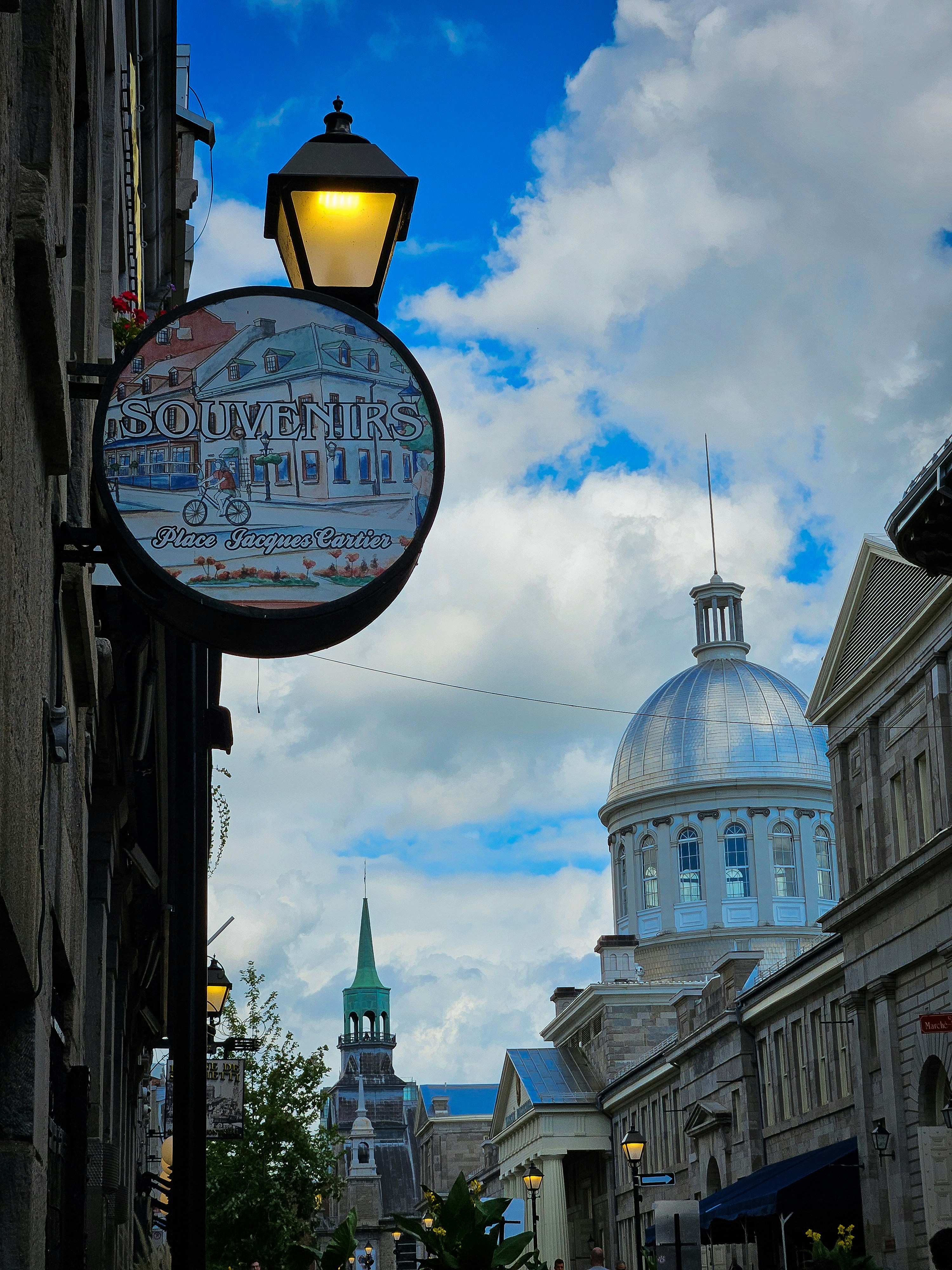 A round sign reading 'Souvenirs' hangs from a lamppost above a cobblestone street, with a neoclassical domed building and a bright blue sky in the background.
