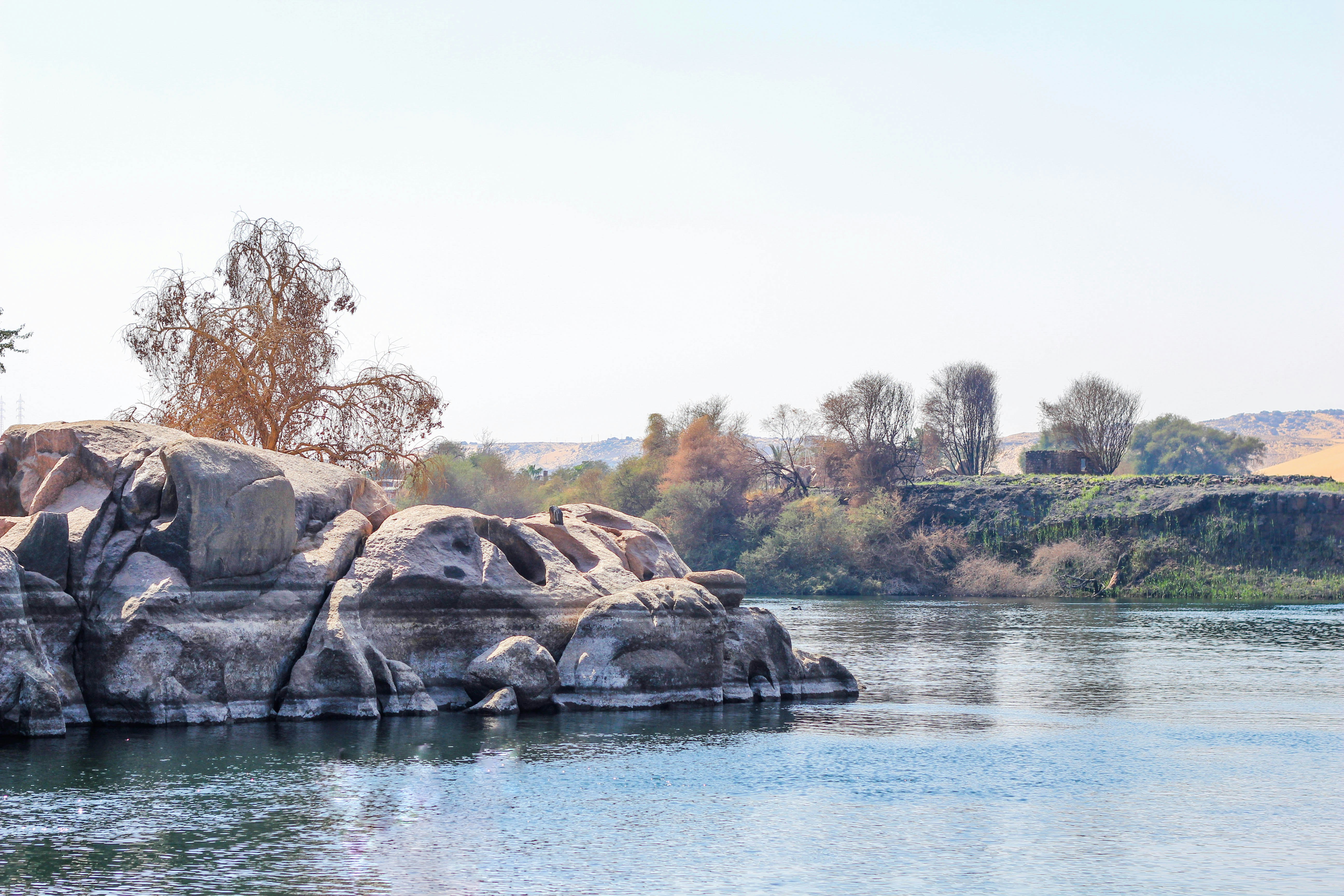 Rocky outcrop along a tranquil river with distant greenery under a clear sky.