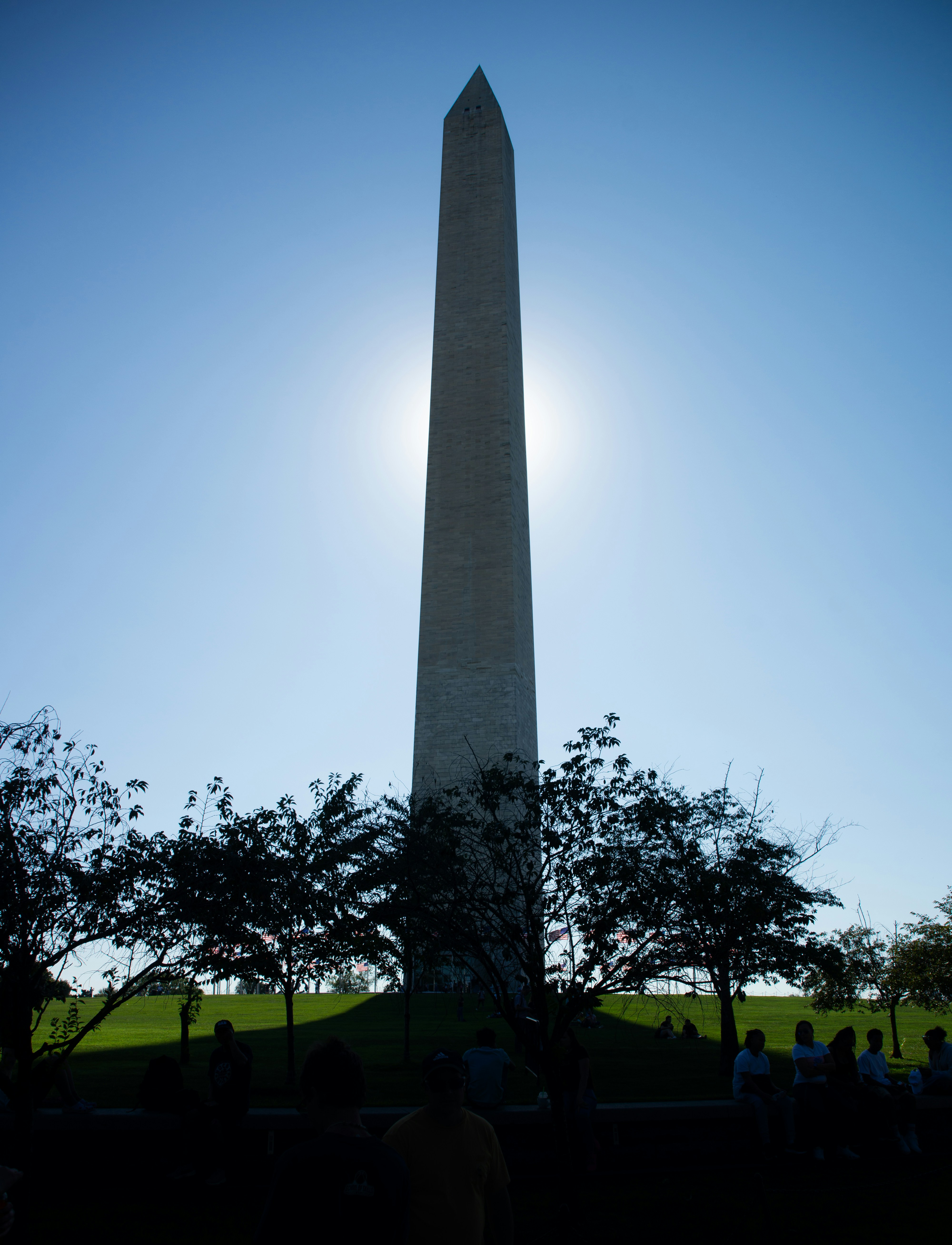 Washington Monument rises against a blue sky with a sun halo, framed by trees and silhouettes of visitors.
