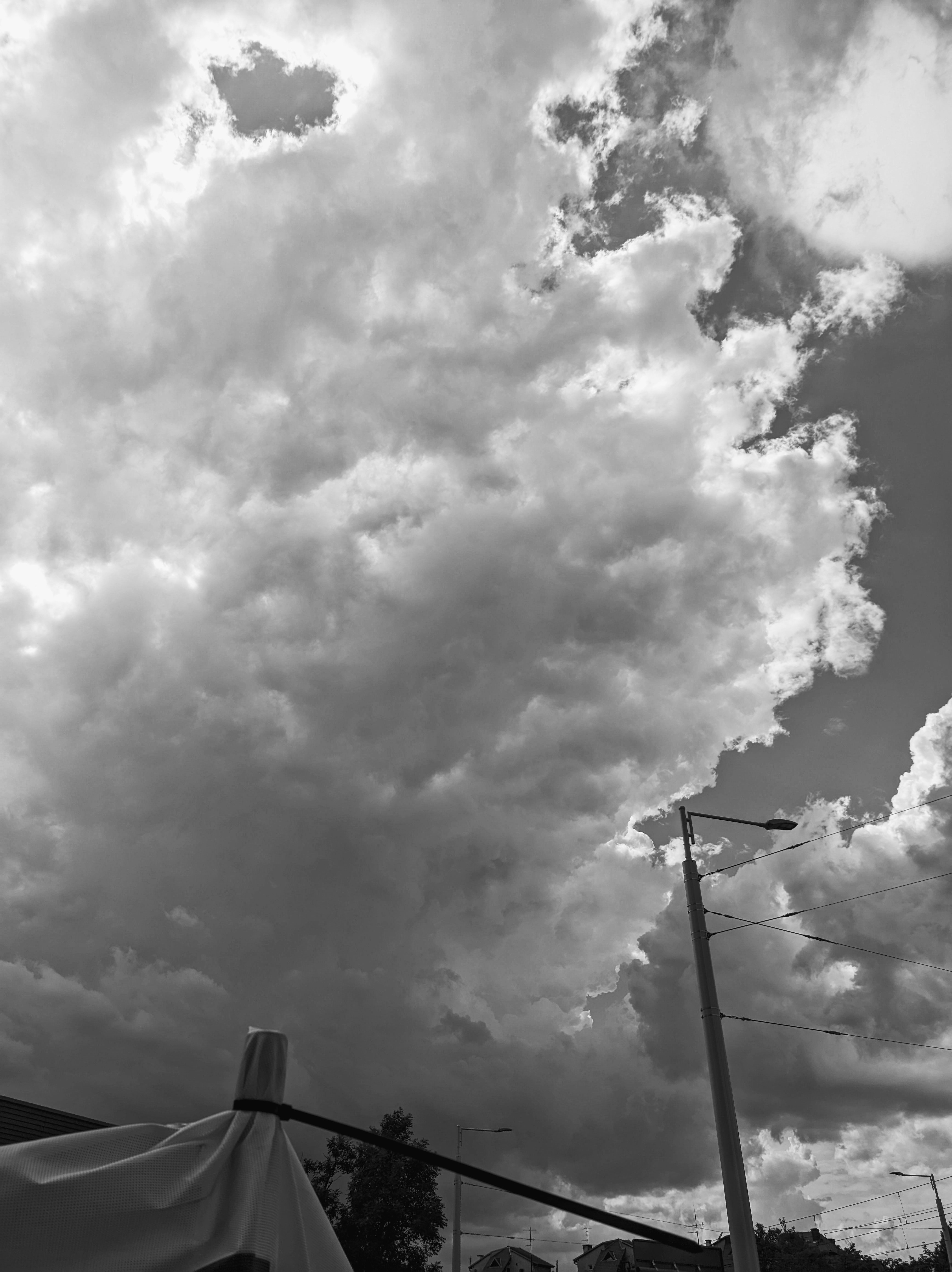 Monochrome photograph of a dramatic cumulus cloudscape looming over a street, with a utility pole and a tarpaulin-covered foreground.