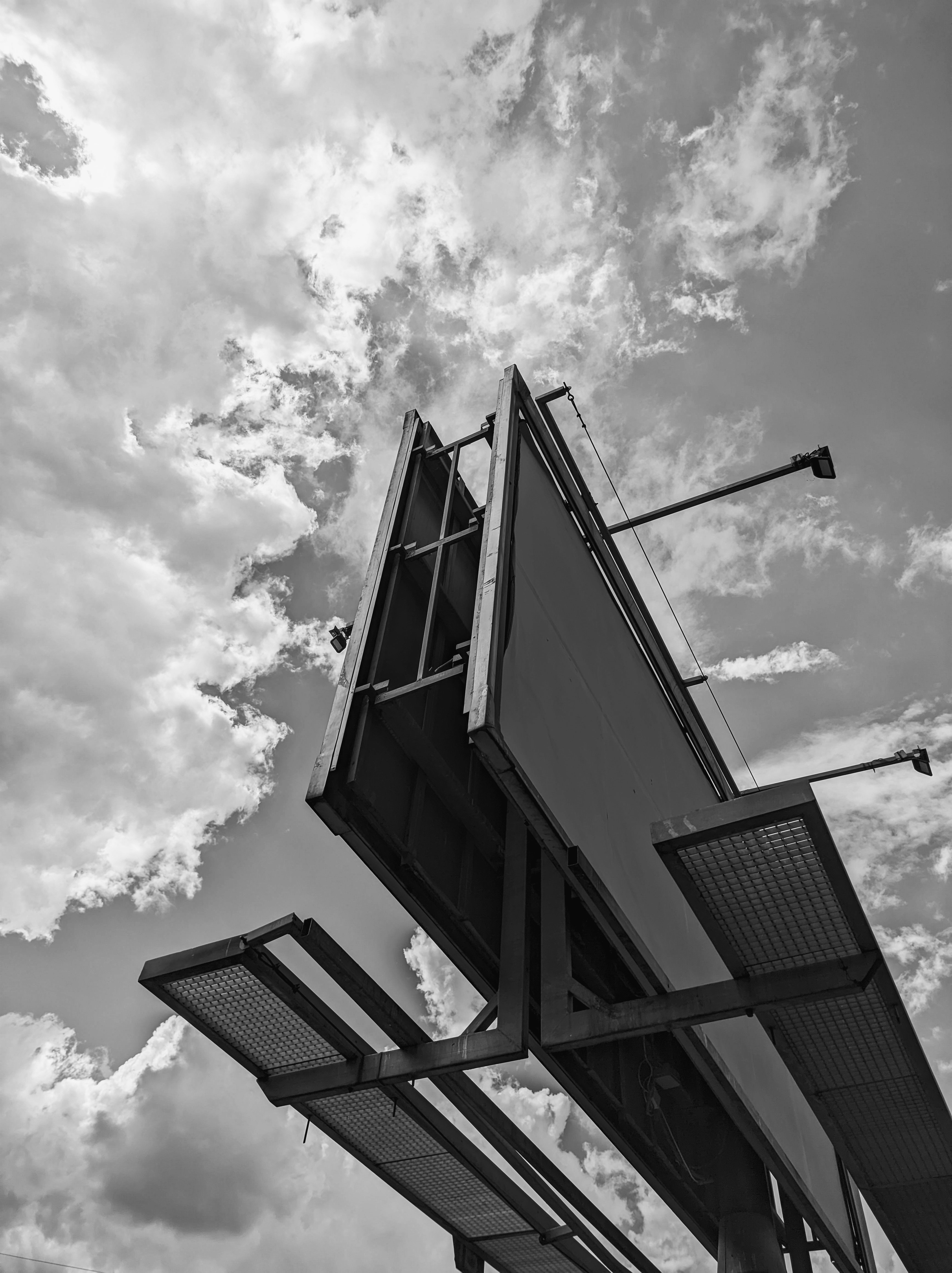 Monochrome shot of a tilted, towering billboard structure against a cloudy sky, emphasizing architectural lines and industrial form.