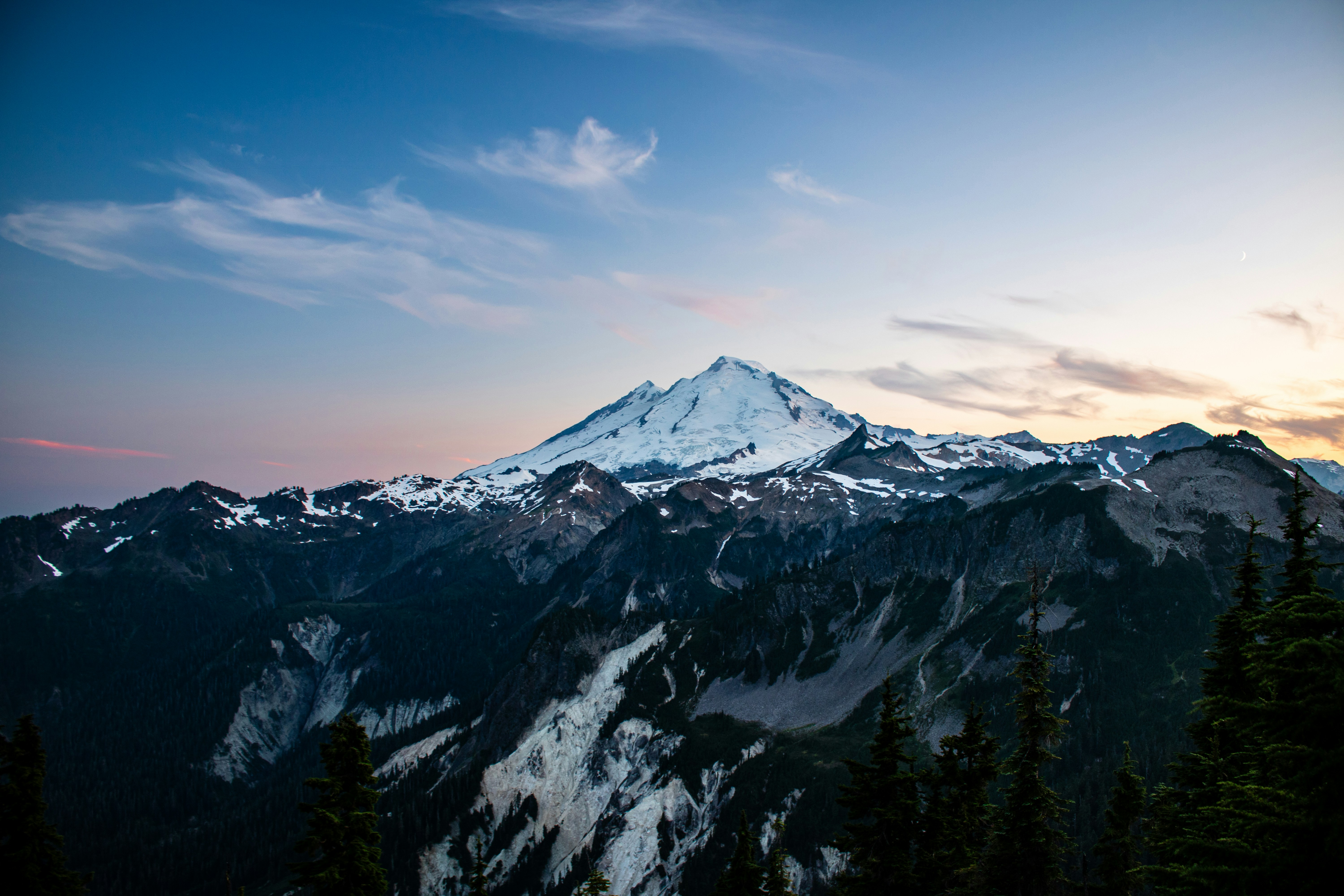 Snow-capped mountain rises above rugged terrain at twilight, framed by evergreen trees. The fading light casts a serene glow over the landscape.