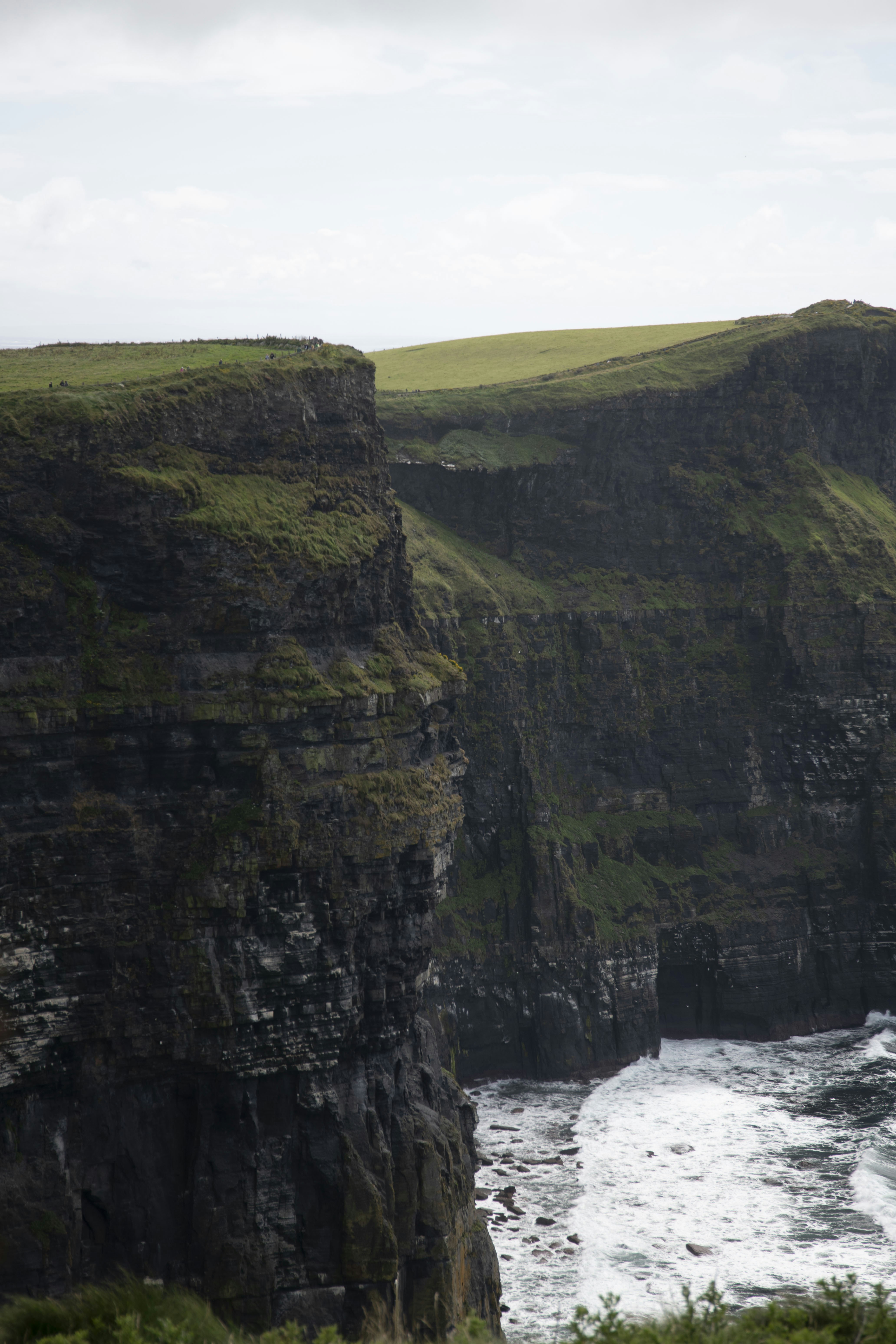 Dramatic cliffs rise above turbulent waves, showcasing the rugged beauty of the coastal landscape. Greenery clings to the cliffside, contrasting with the dark rock formations.