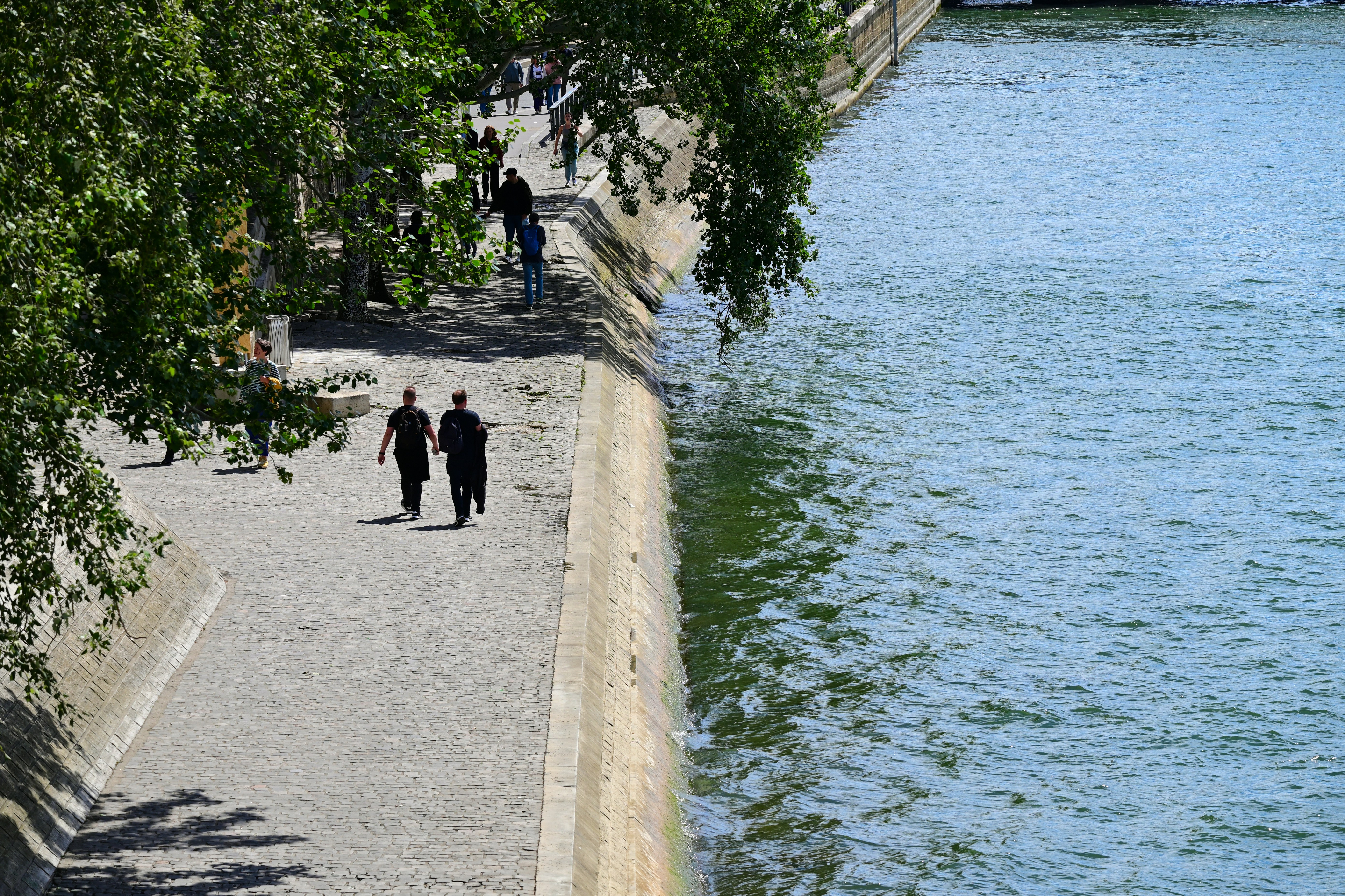 Two figures walking along a riverside path bordered by lush greenery and calm waters. The scene captures a serene afternoon ambiance.