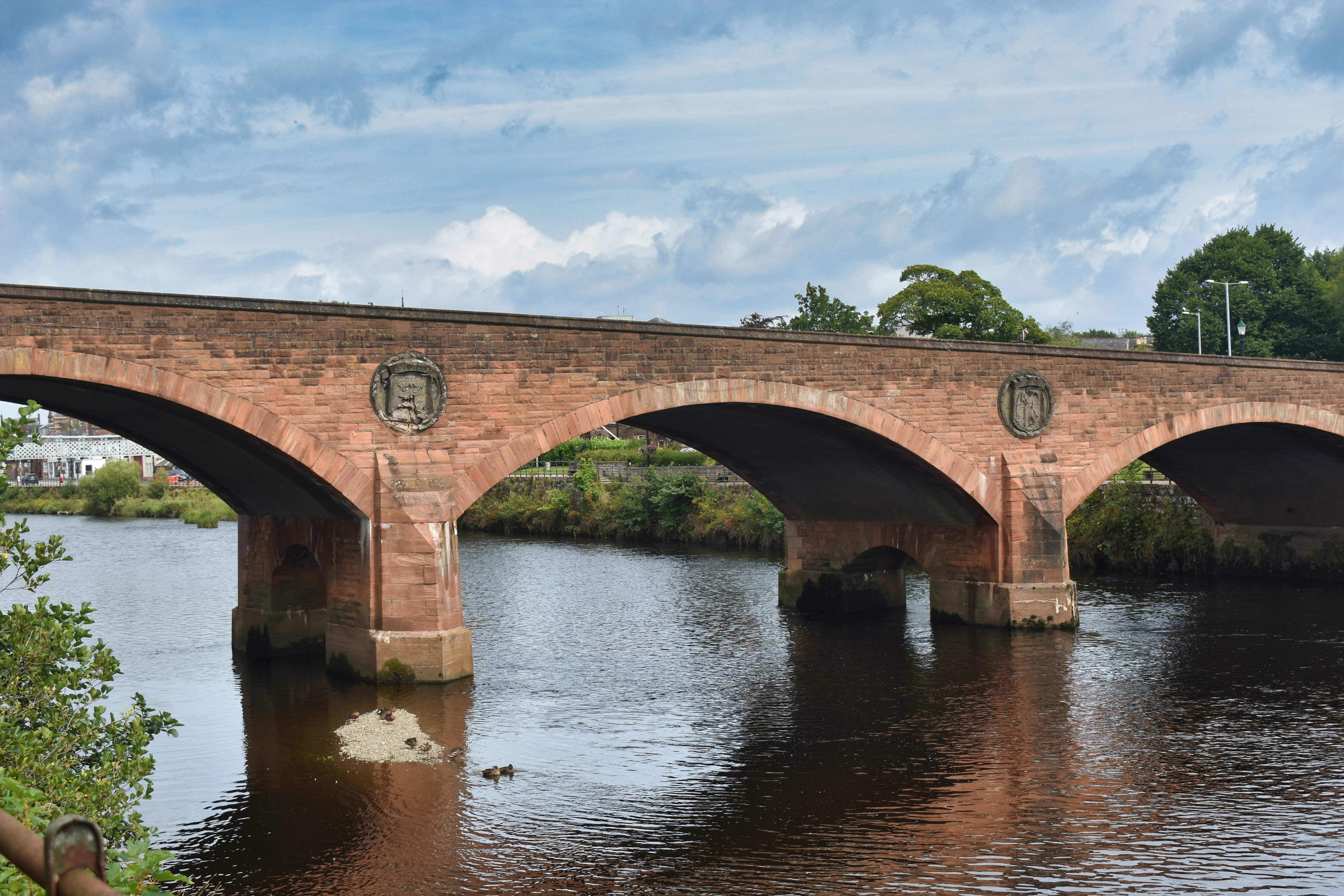 Historic stone bridge spanning a tranquil river, framed by lush greenery and a cloudy sky.