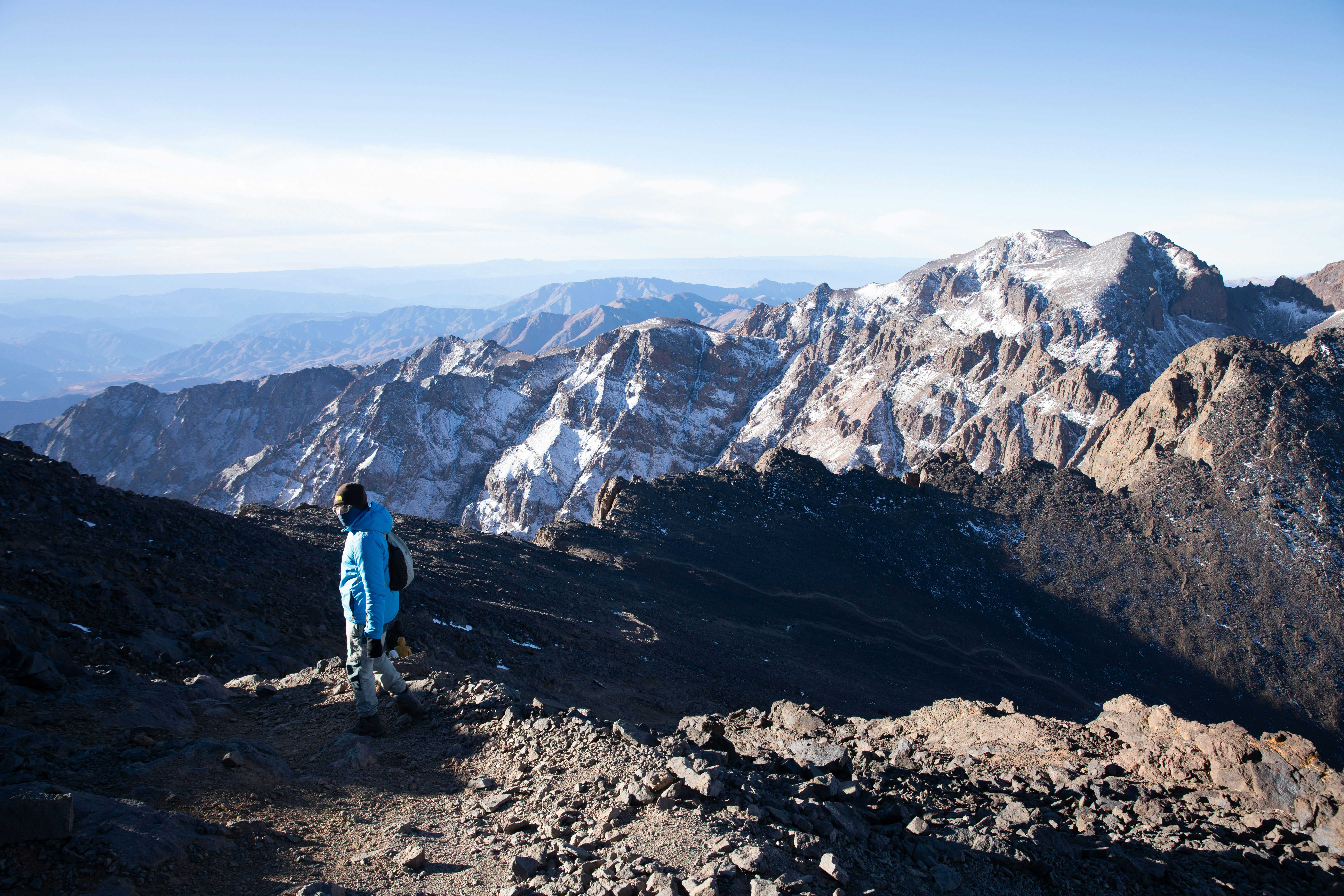 Hiker in blue jacket traversing rocky mountain ridge with expansive snow-capped peaks in the background.