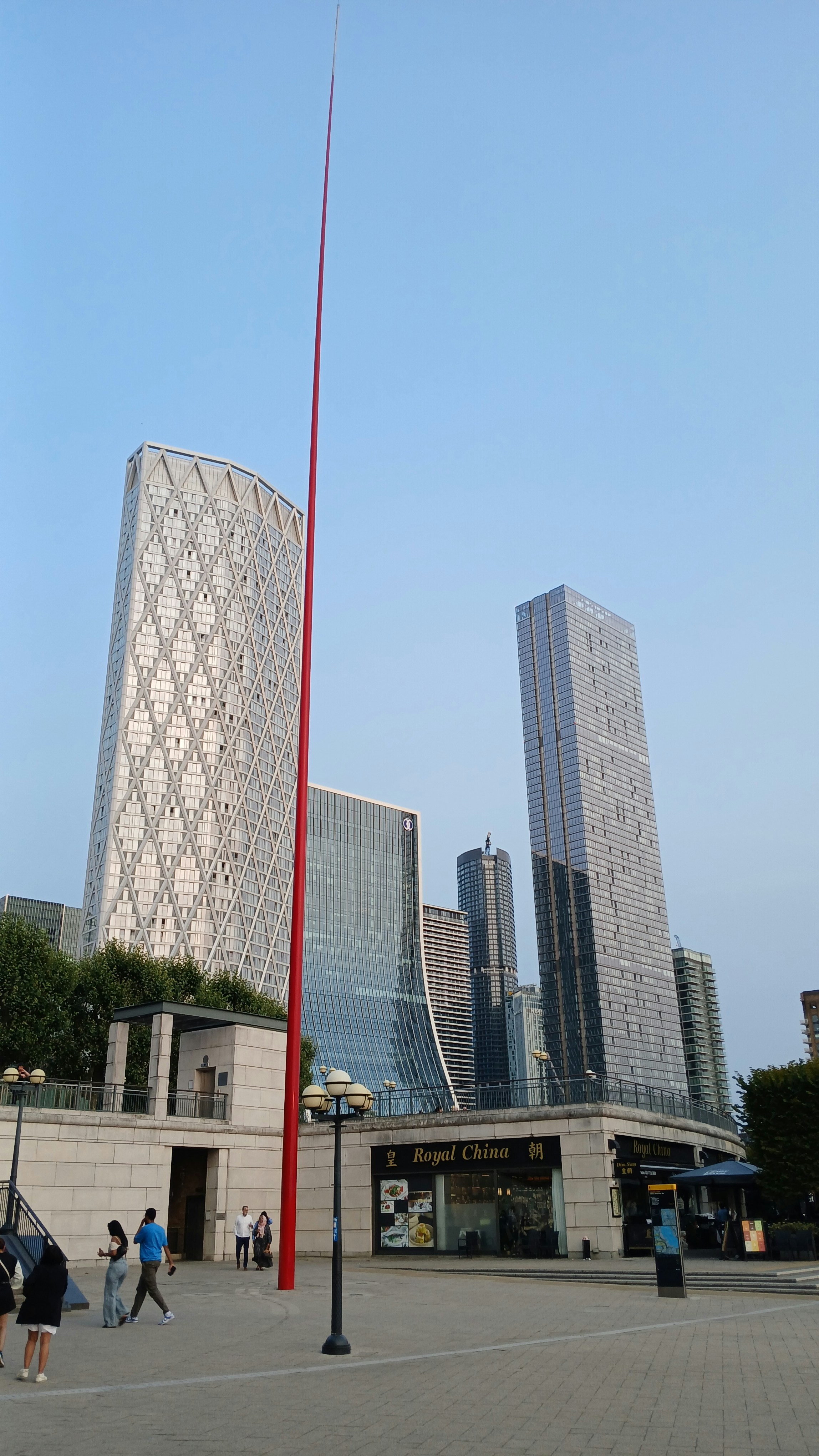 Tall crimson mast rises beside sleek glass towers in a sunlit urban plaza.